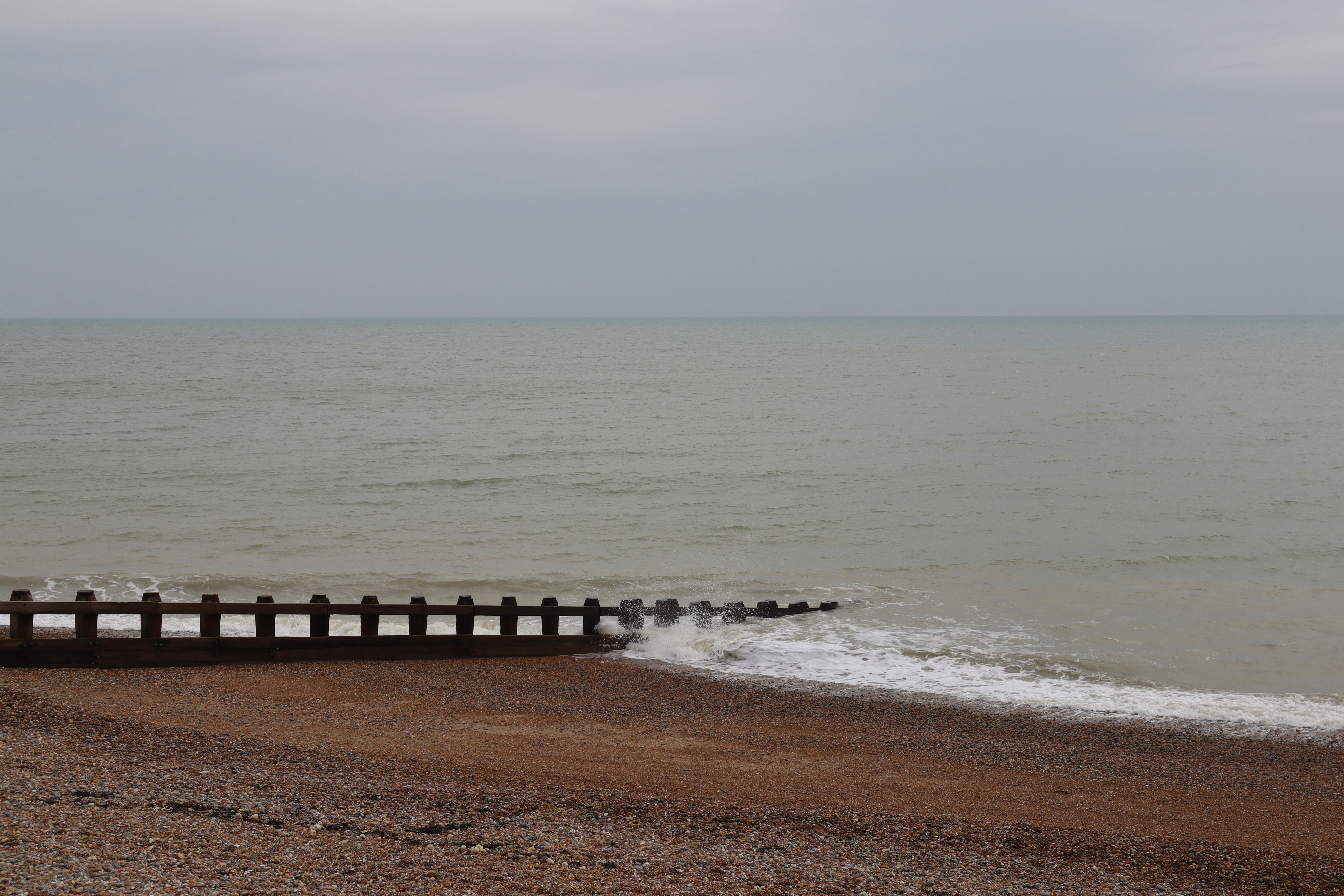 Calm sea meets a pebbled beach with a wooden breakwater under cloudy skies.