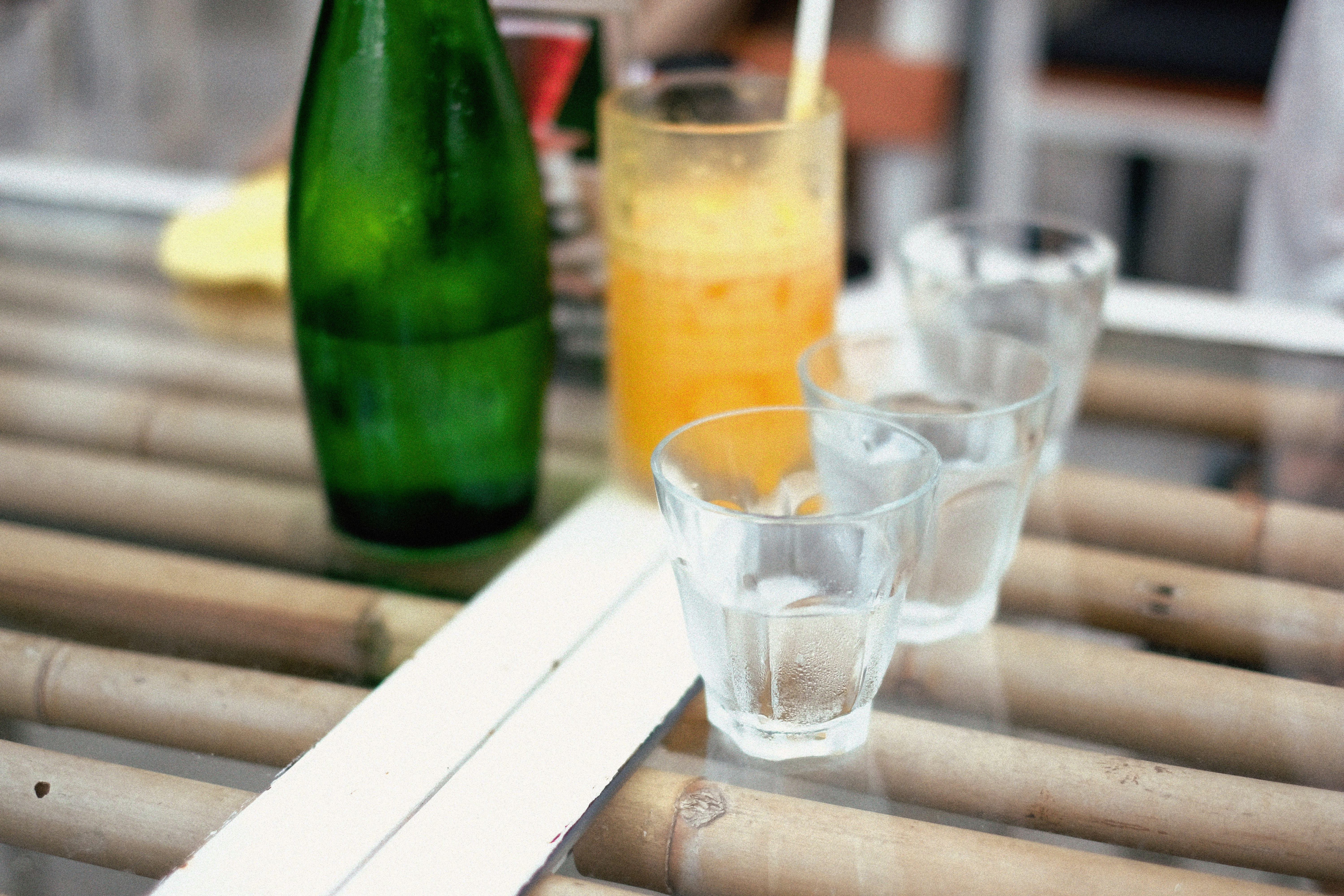 A green bottle and a vibrant orange drink sit atop a bamboo table, surrounded by empty glasses, suggesting a social gathering or a moment of relaxation.
