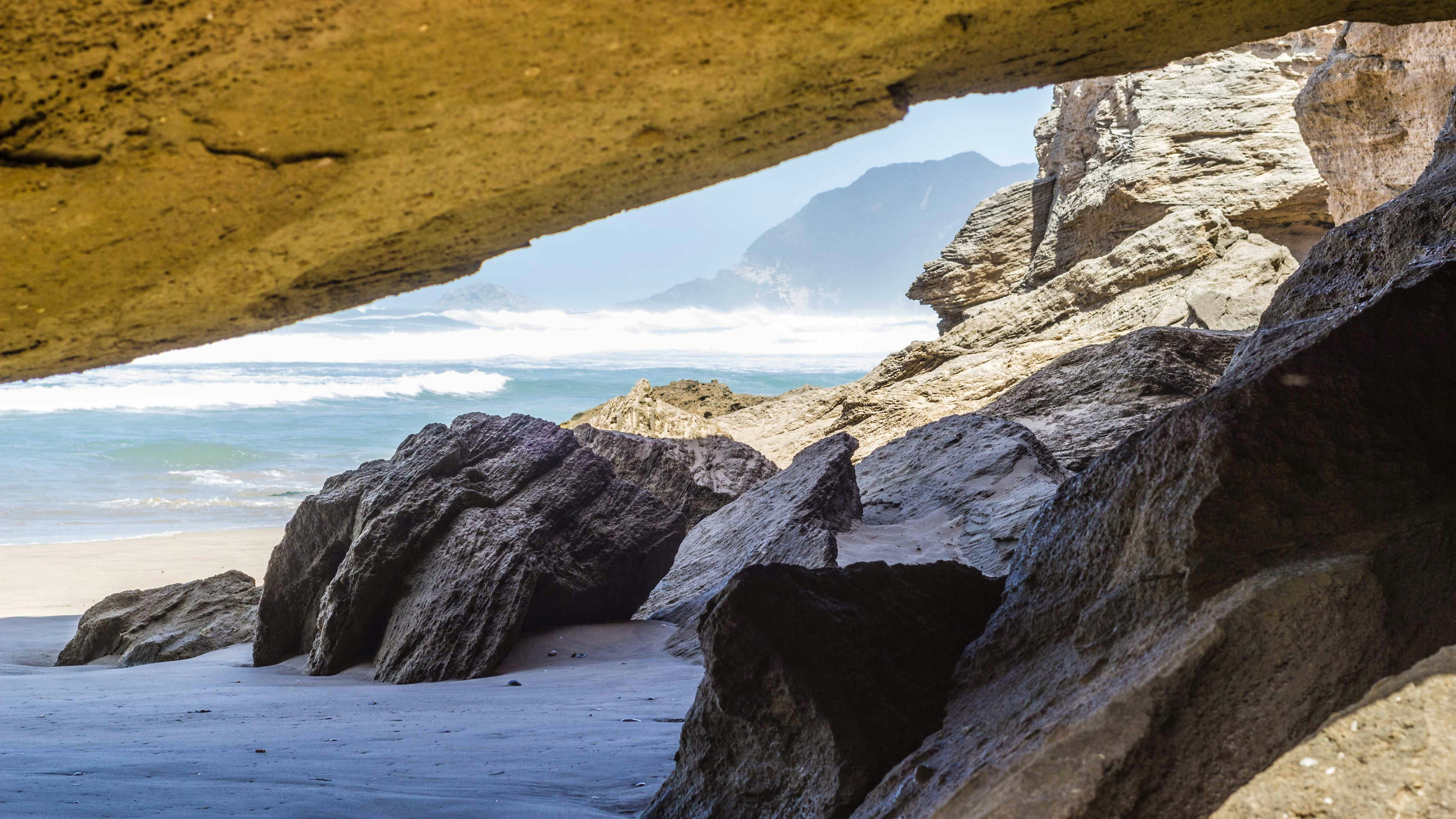 Rocky beach scene framed by a natural archway, revealing the ocean and distant mountains. The interplay of light and shadow adds depth to the coastal landscape.