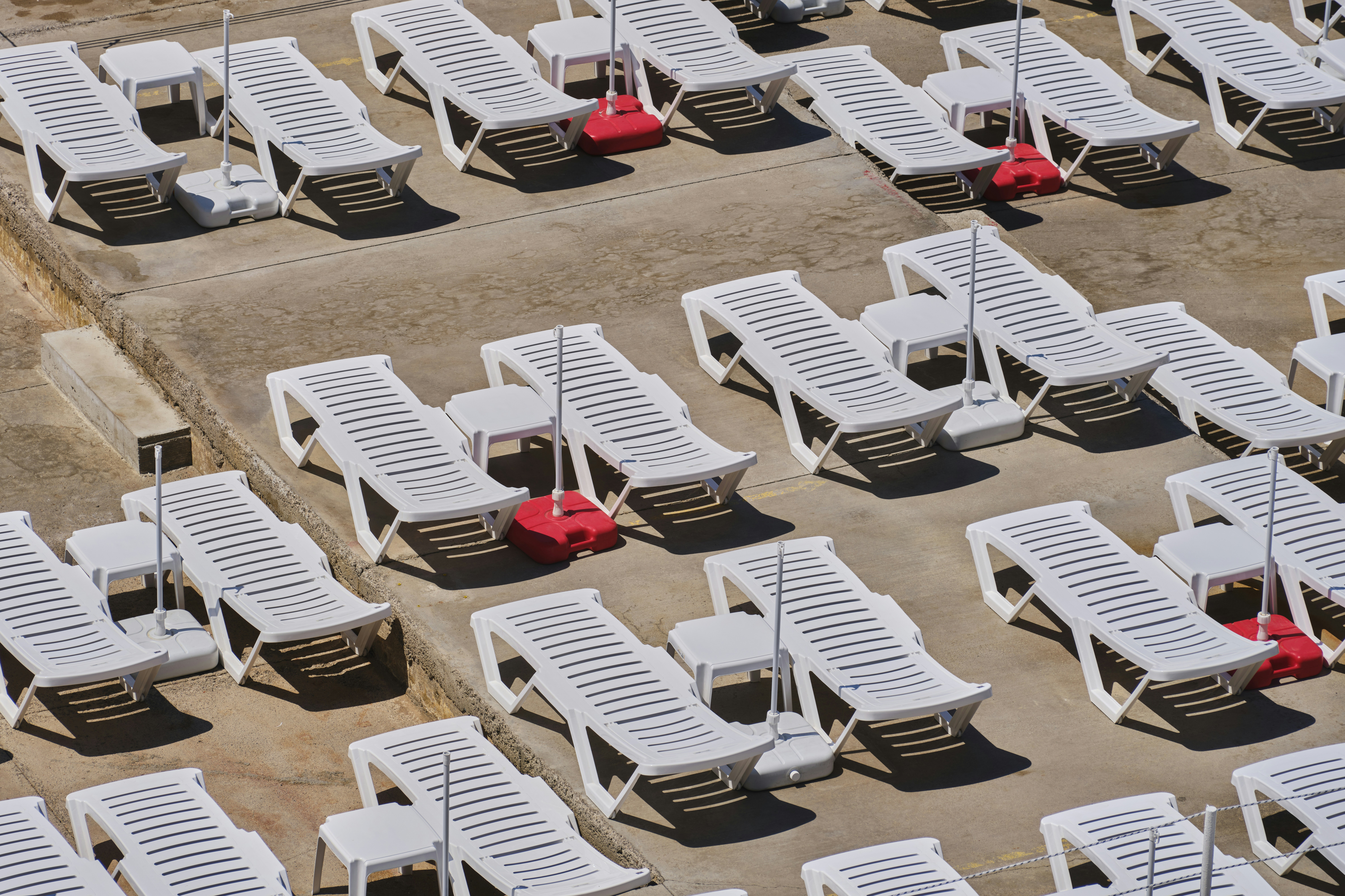 Aerial photograph of neatly arranged white sun loungers with a few red cushions, spread across a sunlit poolside deck at a beach club.