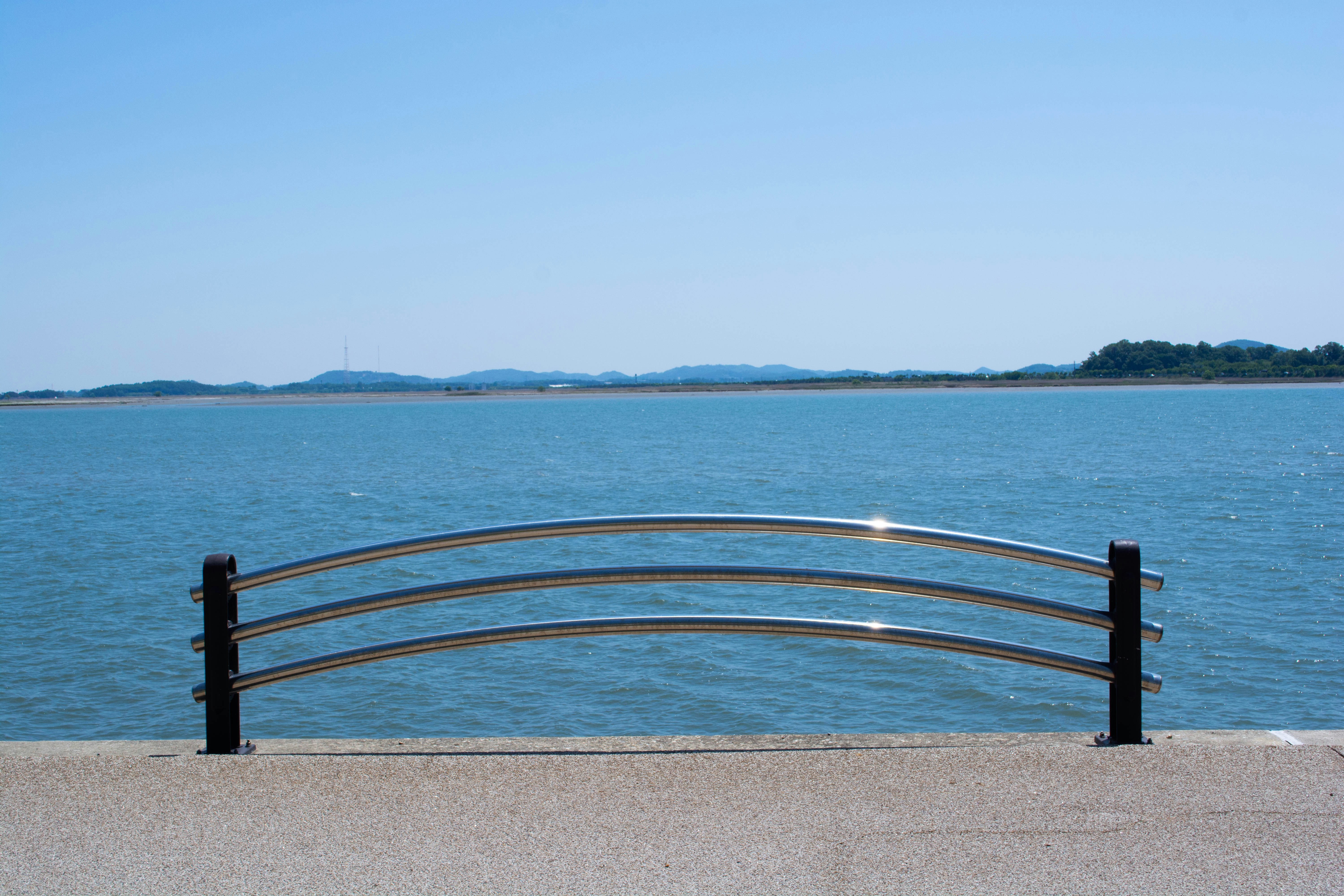 Curved metal railing overlooking a tranquil body of water with distant hills under a clear blue sky.