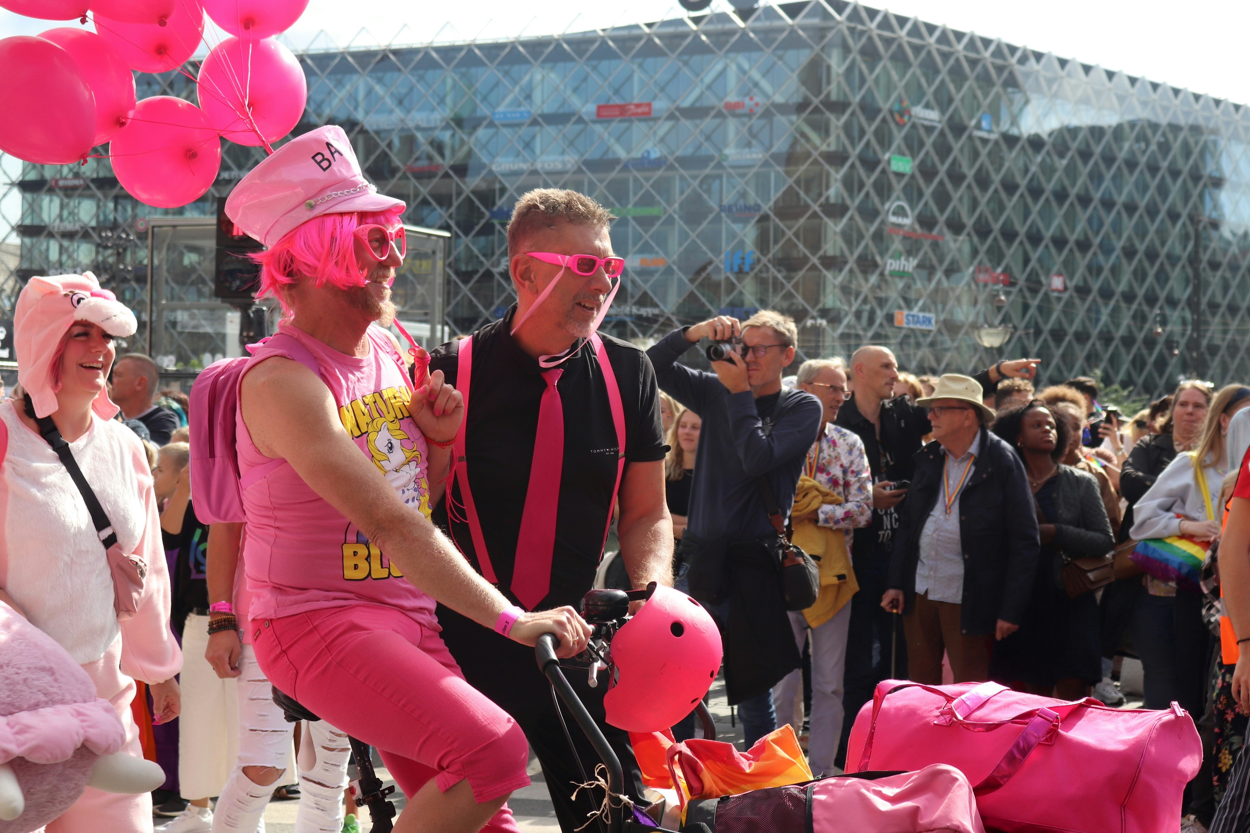Two men dressed in vibrant pink attire ride a bicycle, surrounded by a lively crowd at a festive event. Balloons and colorful outfits add to the celebratory atmosphere.