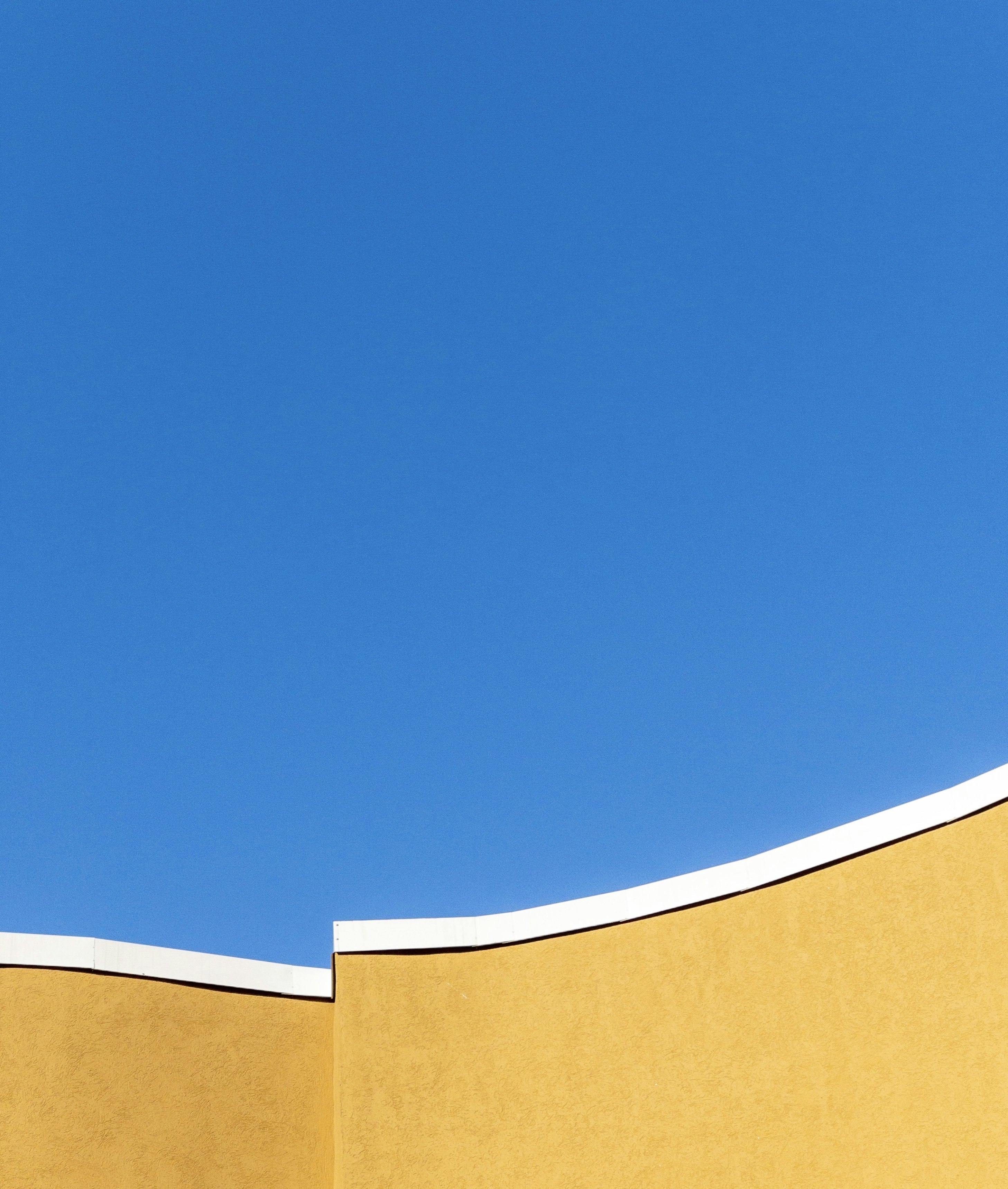 A minimalist architectural detail featuring a yellow wall with a white edge, set against a clear blue sky.