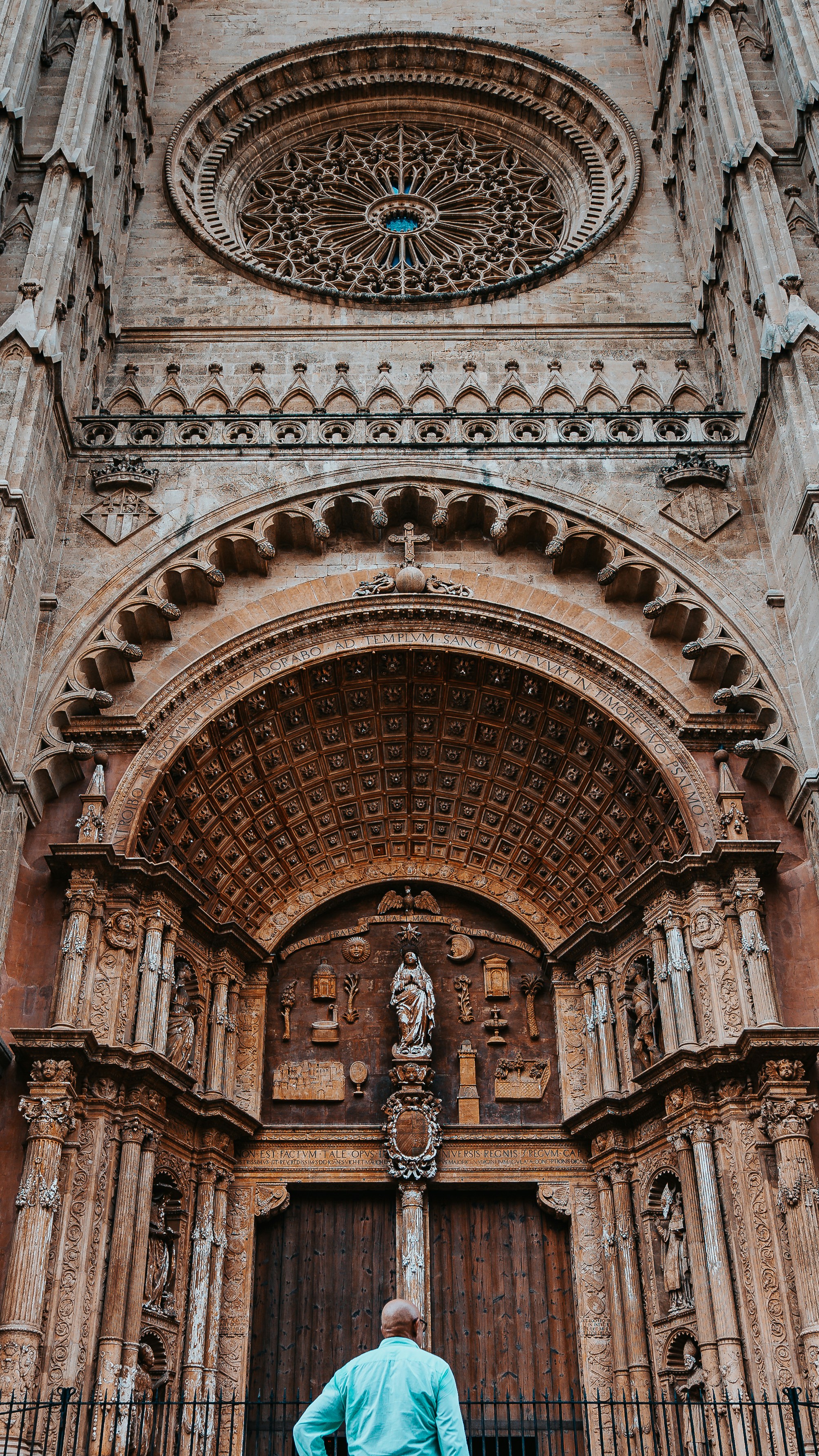 Intricate facade of a historical building featuring ornate sculptures and a circular window, with a figure standing in awe at the entrance.