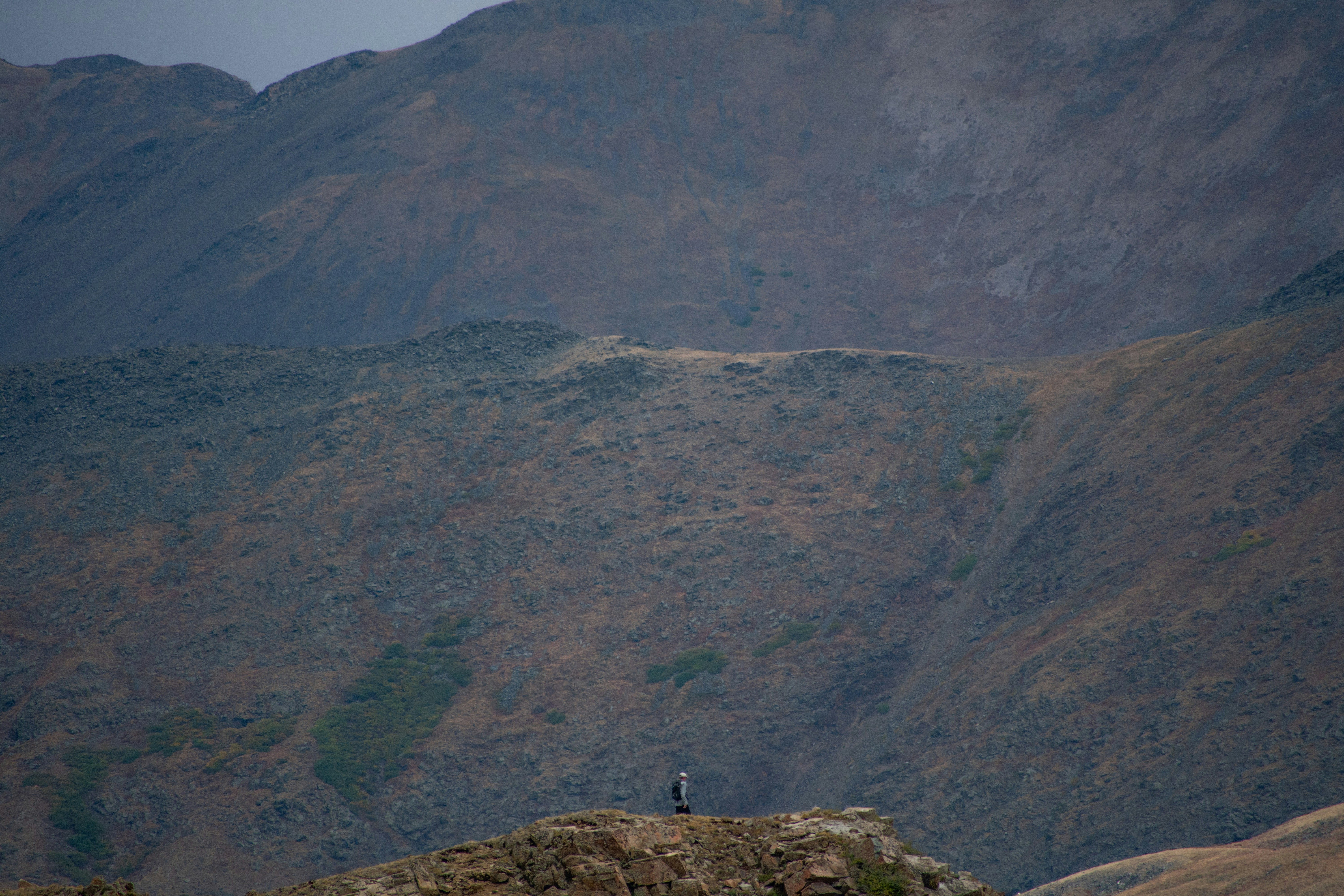 A lone individual stands atop a rocky outcrop, surrounded by expansive mountain ranges in muted earthy tones.
