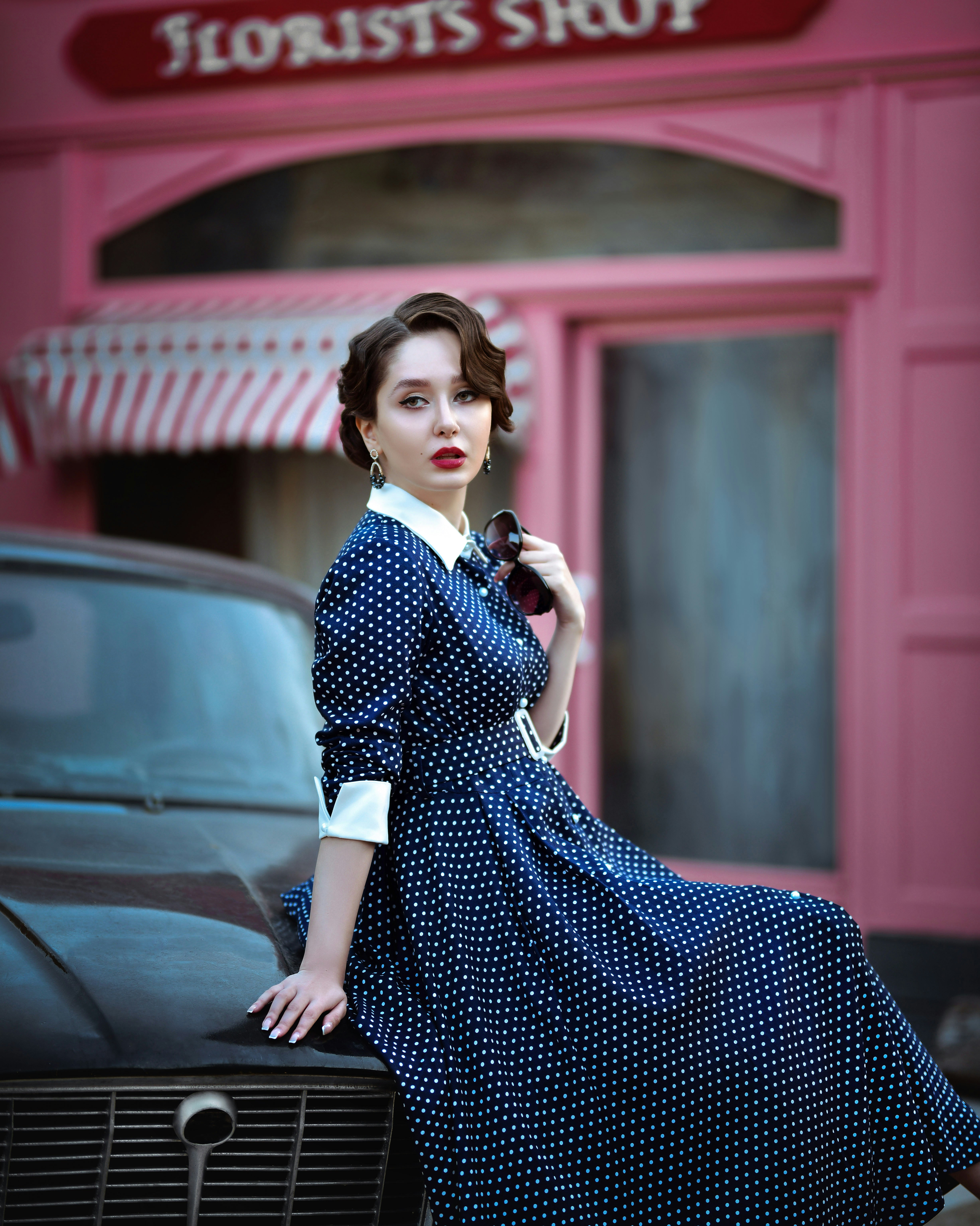 A woman in a polka dot dress poses gracefully on a classic car, with a charming florist shop backdrop. The scene evokes a nostalgic atmosphere.