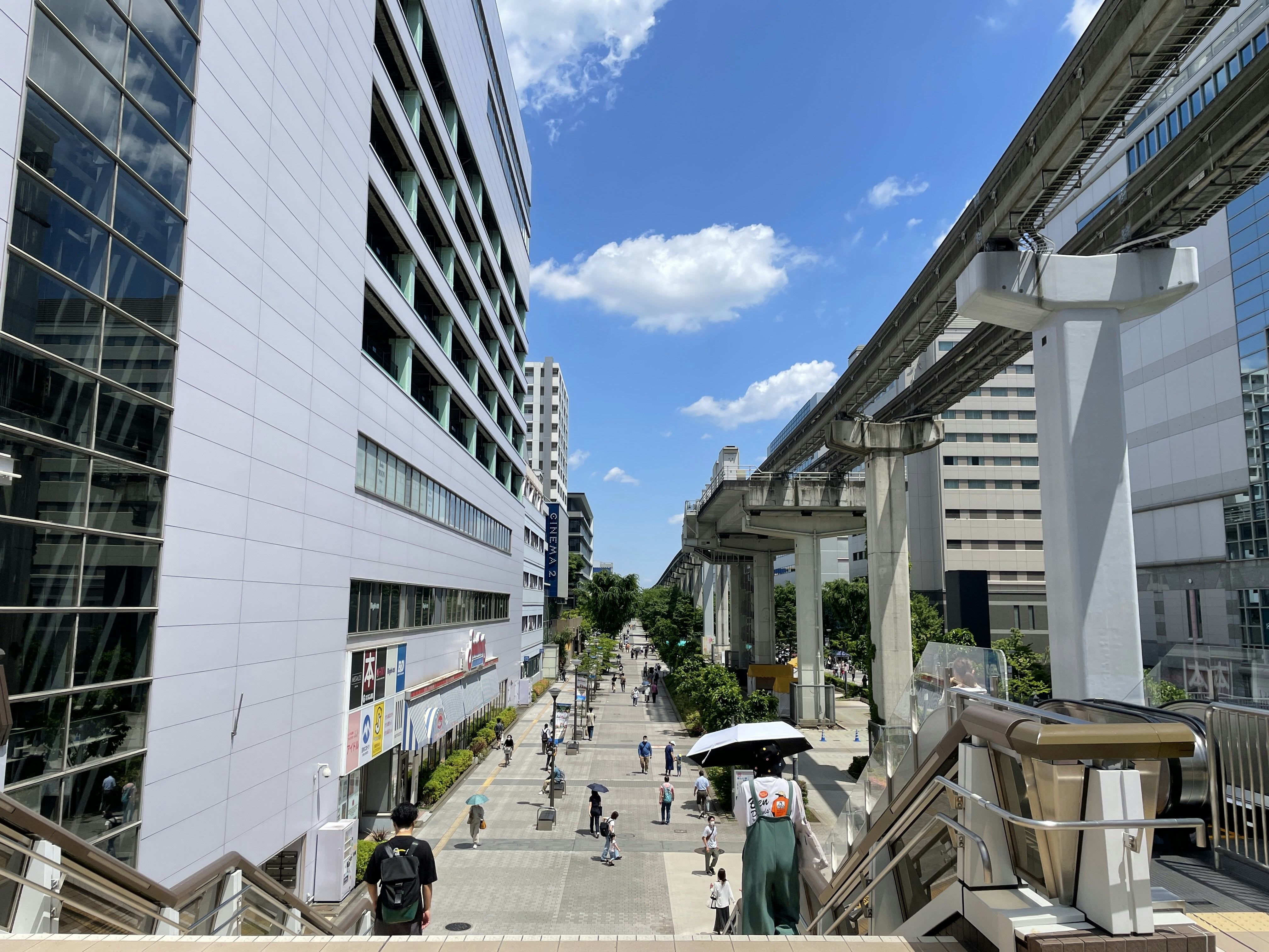 A bustling urban scene with pedestrians walking along a wide path, framed by modern architecture and an elevated train track above.