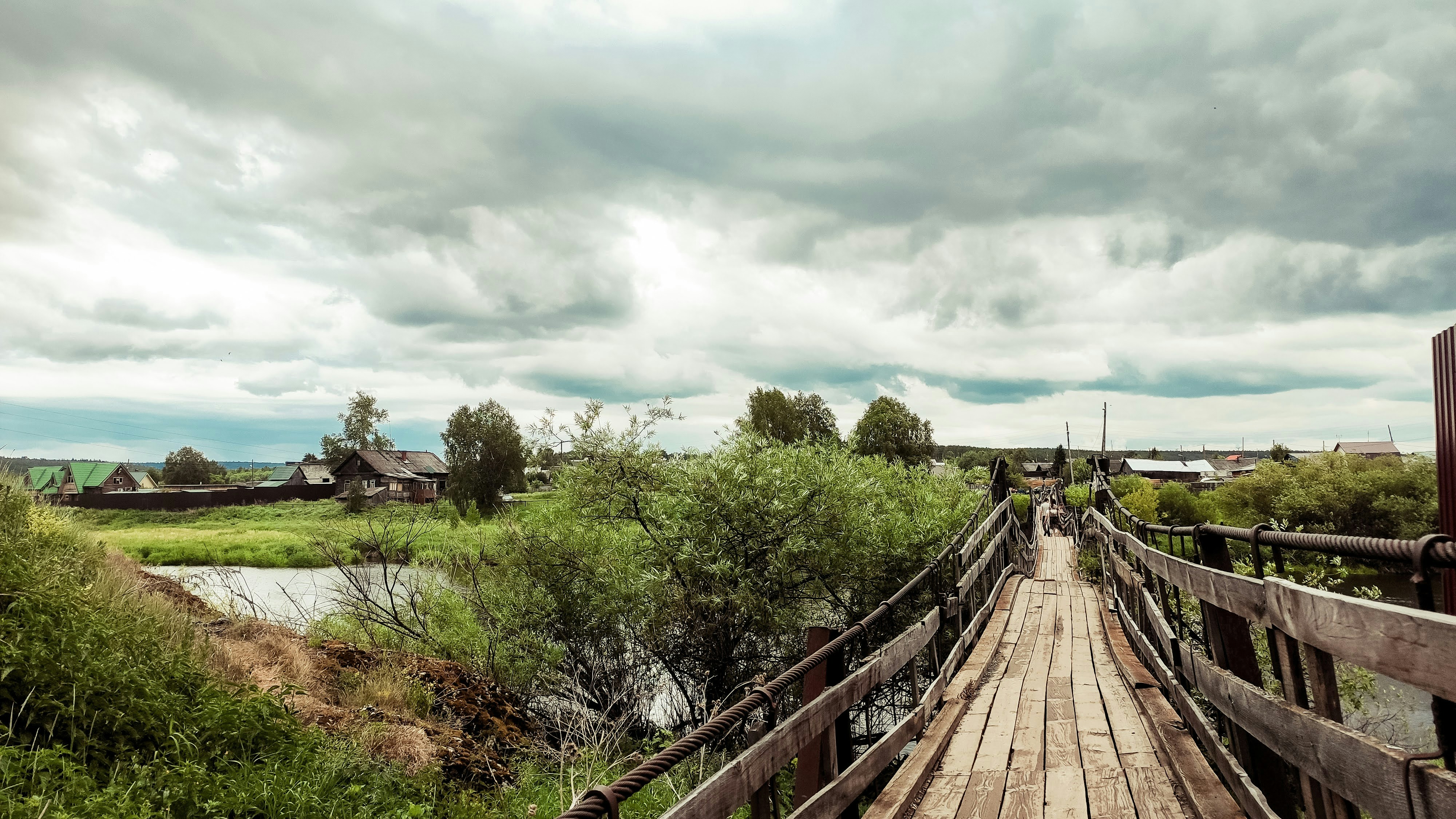 Wooden bridge leading across a serene river, surrounded by lush greenery and traditional houses under a cloudy sky.