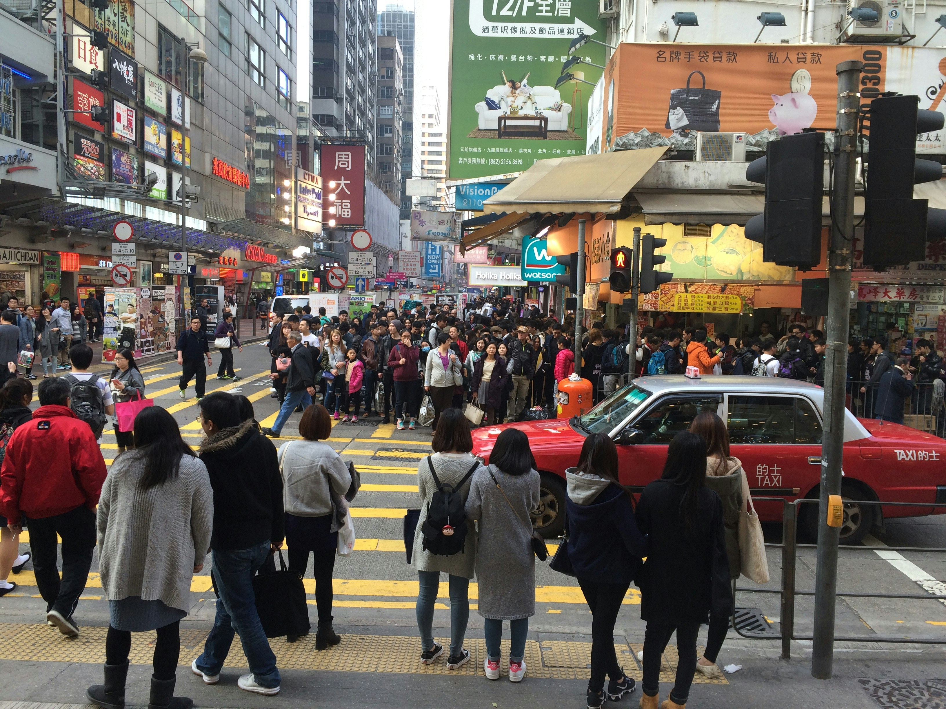 Crowd of pedestrians waiting to cross a busy street in a vibrant urban setting, with a red taxi in the foreground and colorful shop signs lining the background.
