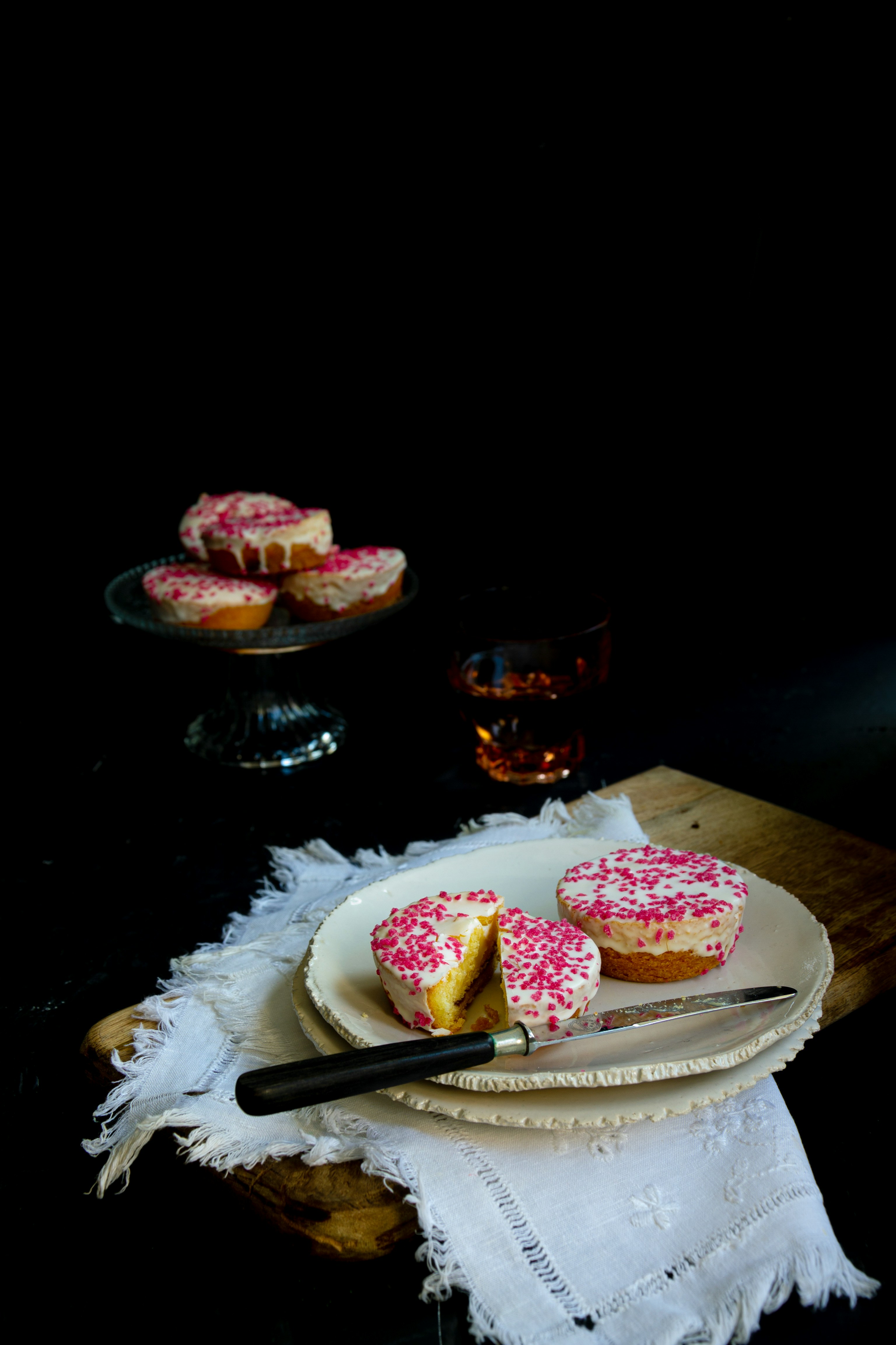 Pink sprinkle-covered pastries on a white plate with a knife, set on a wooden board with a dark background.