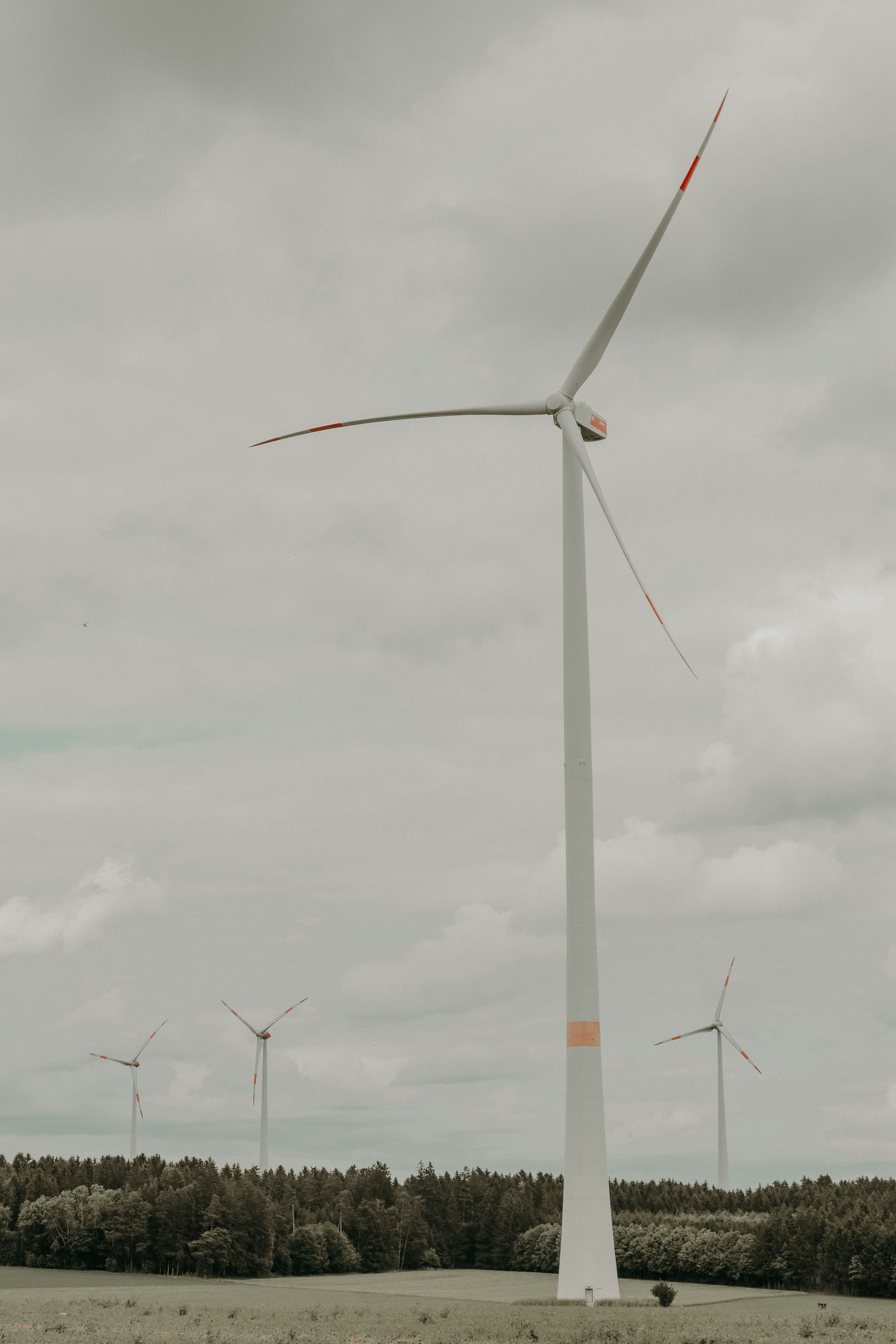 A towering wind turbine stands prominently against a backdrop of cloudy skies and distant turbines, symbolizing renewable energy. The lush landscape below contrasts with the mechanical elegance above.