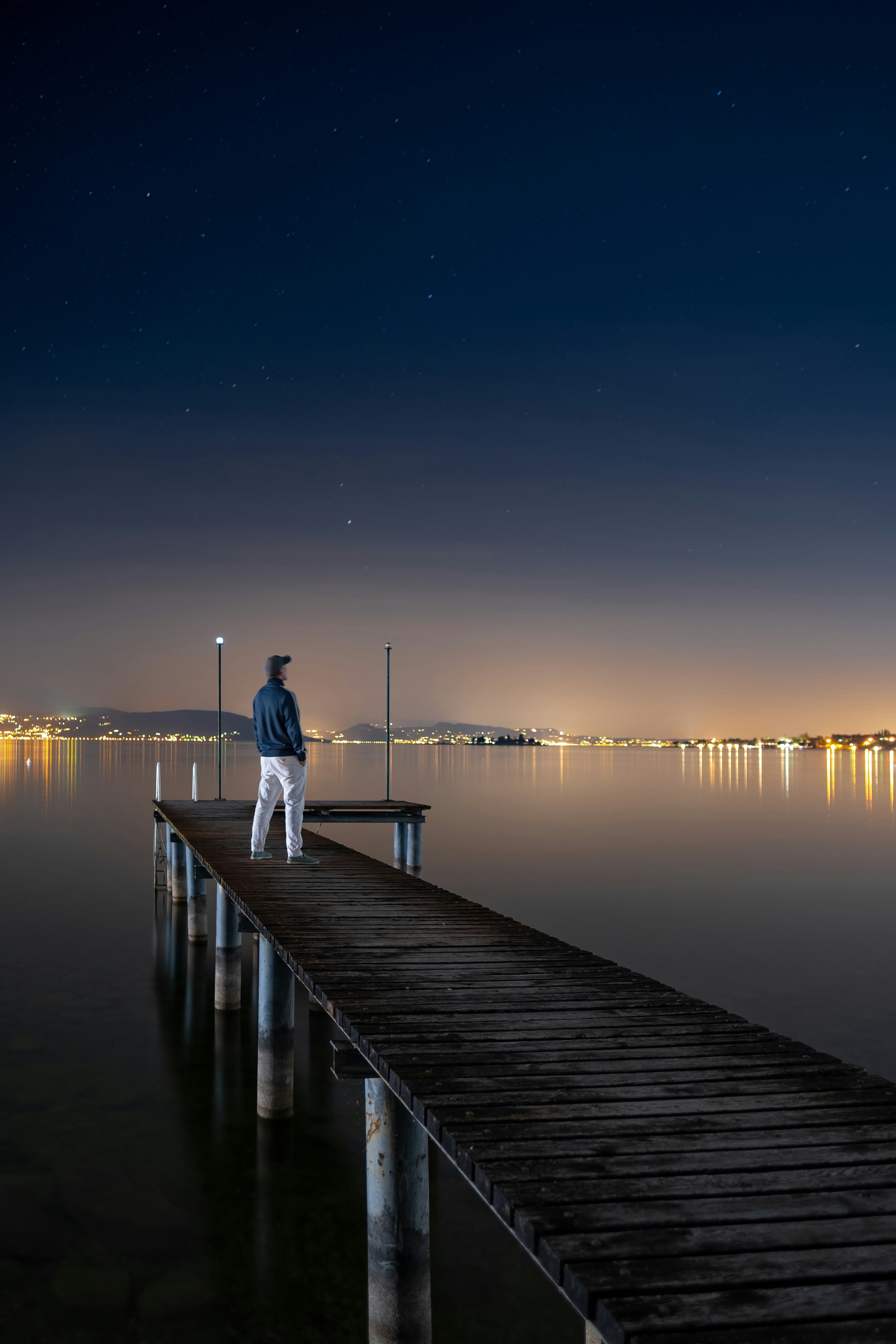 Person standing on a wooden pier overlooking calm waters under a starry night sky.
