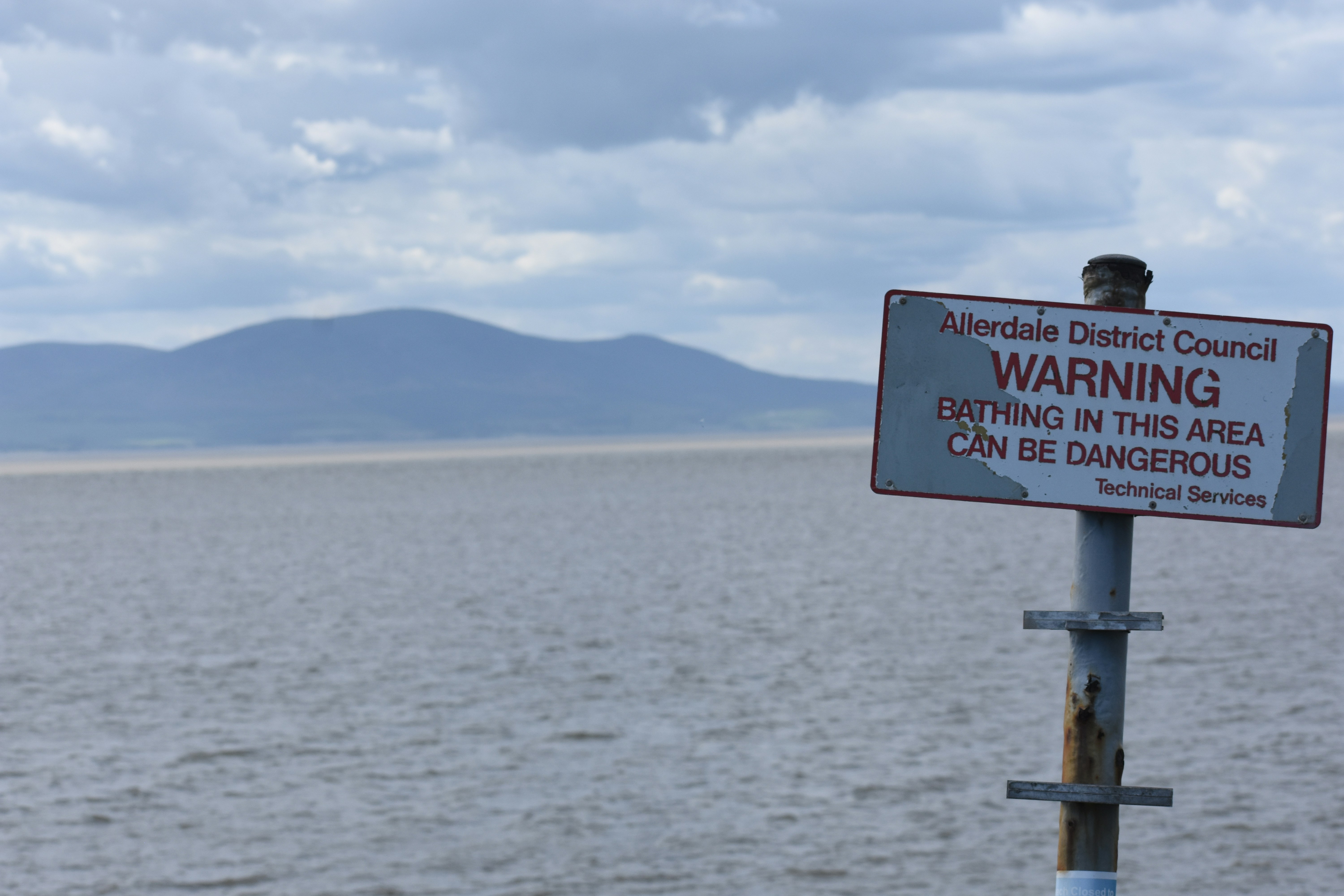 Warning Sign at the Edge of a Glassy Lake Under Cloudy Hills