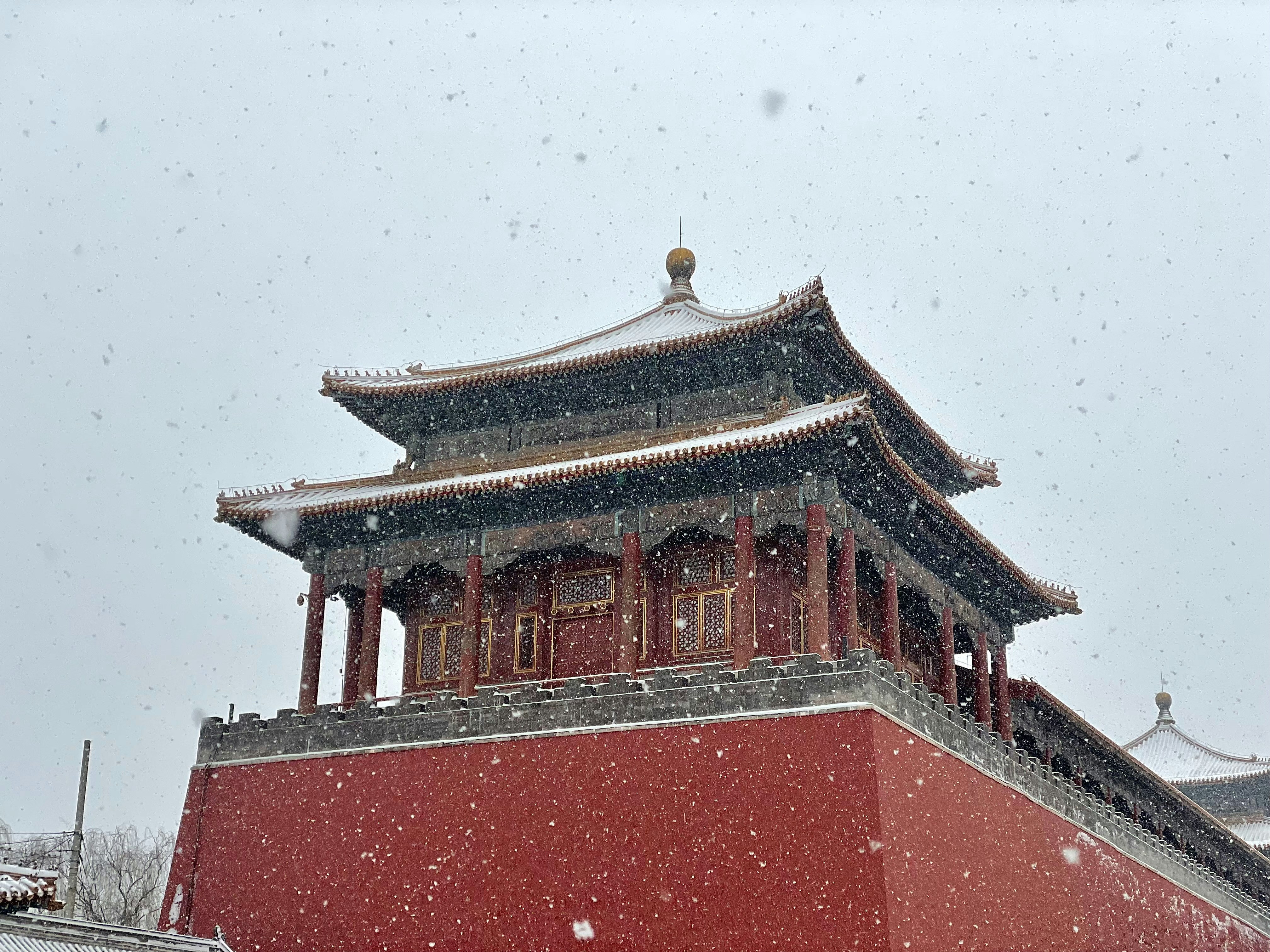 Traditional Chinese pavilion adorned with intricate details, partially obscured by falling snowflakes. A serene winter scene unfolds around the historic architecture.