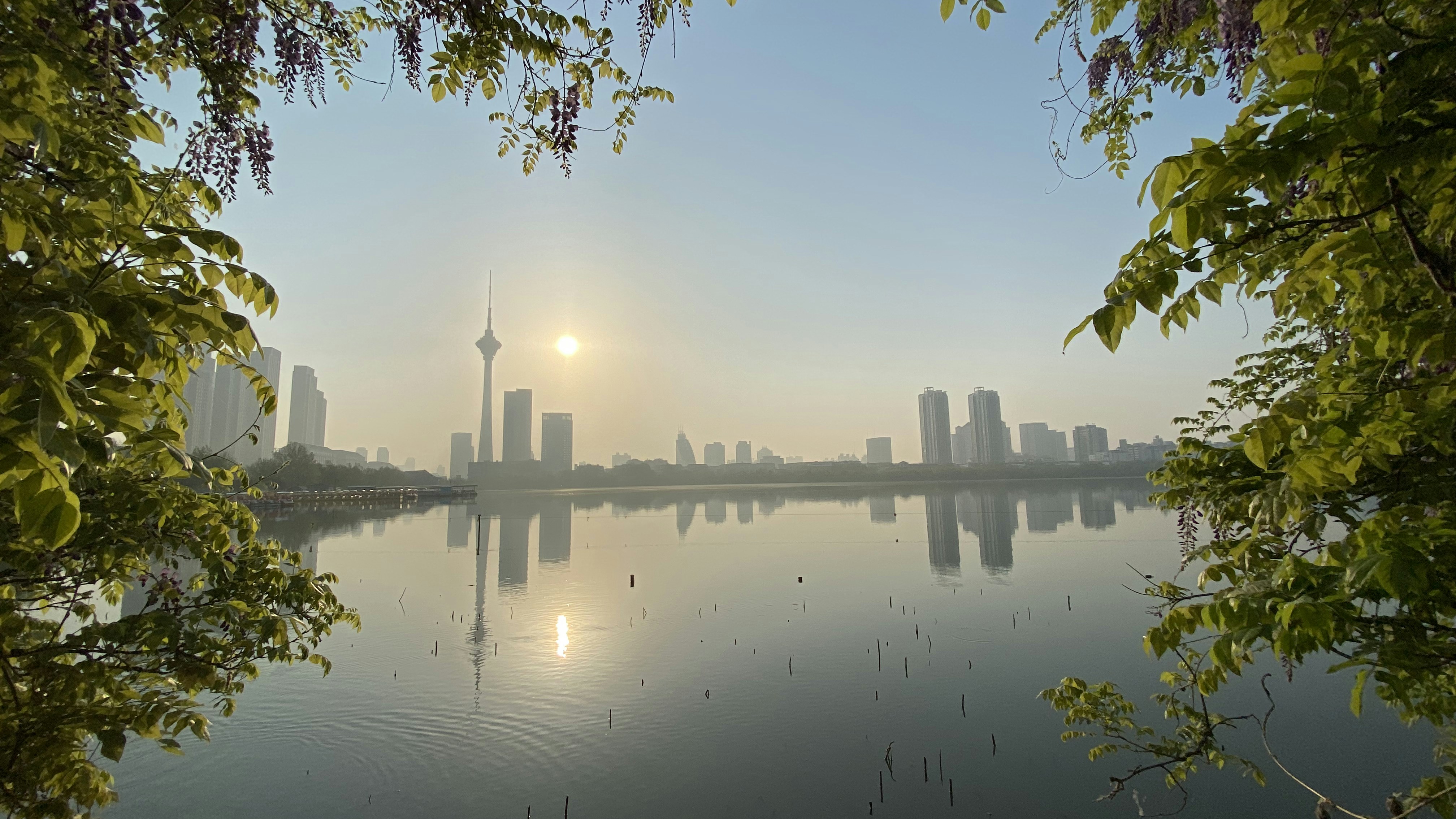 City skyline mirrored in calm waters framed by lush greenery at sunrise.