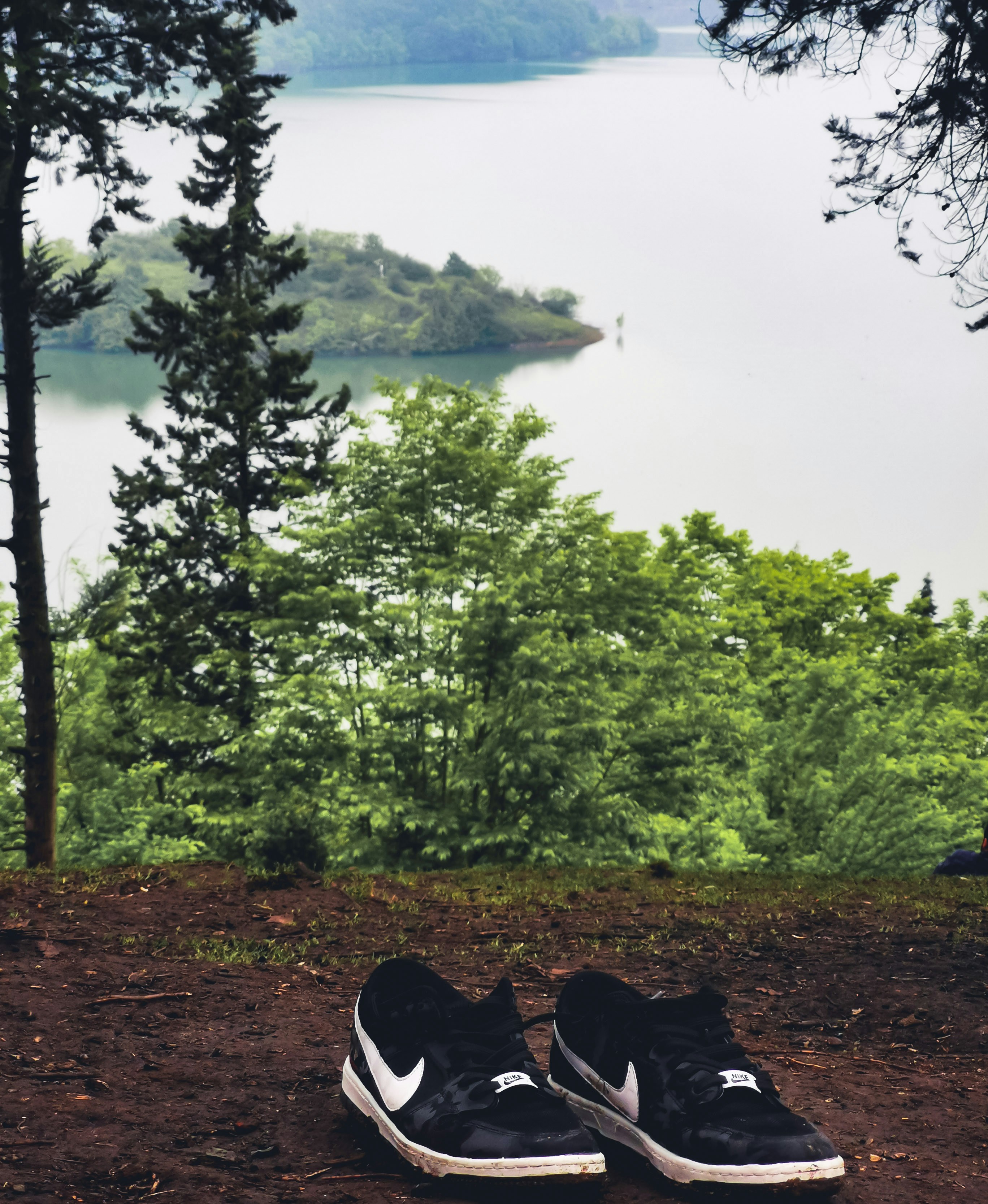A pair of black sneakers rests on the dirt foreground as lush trees frame a calm lake beyond.