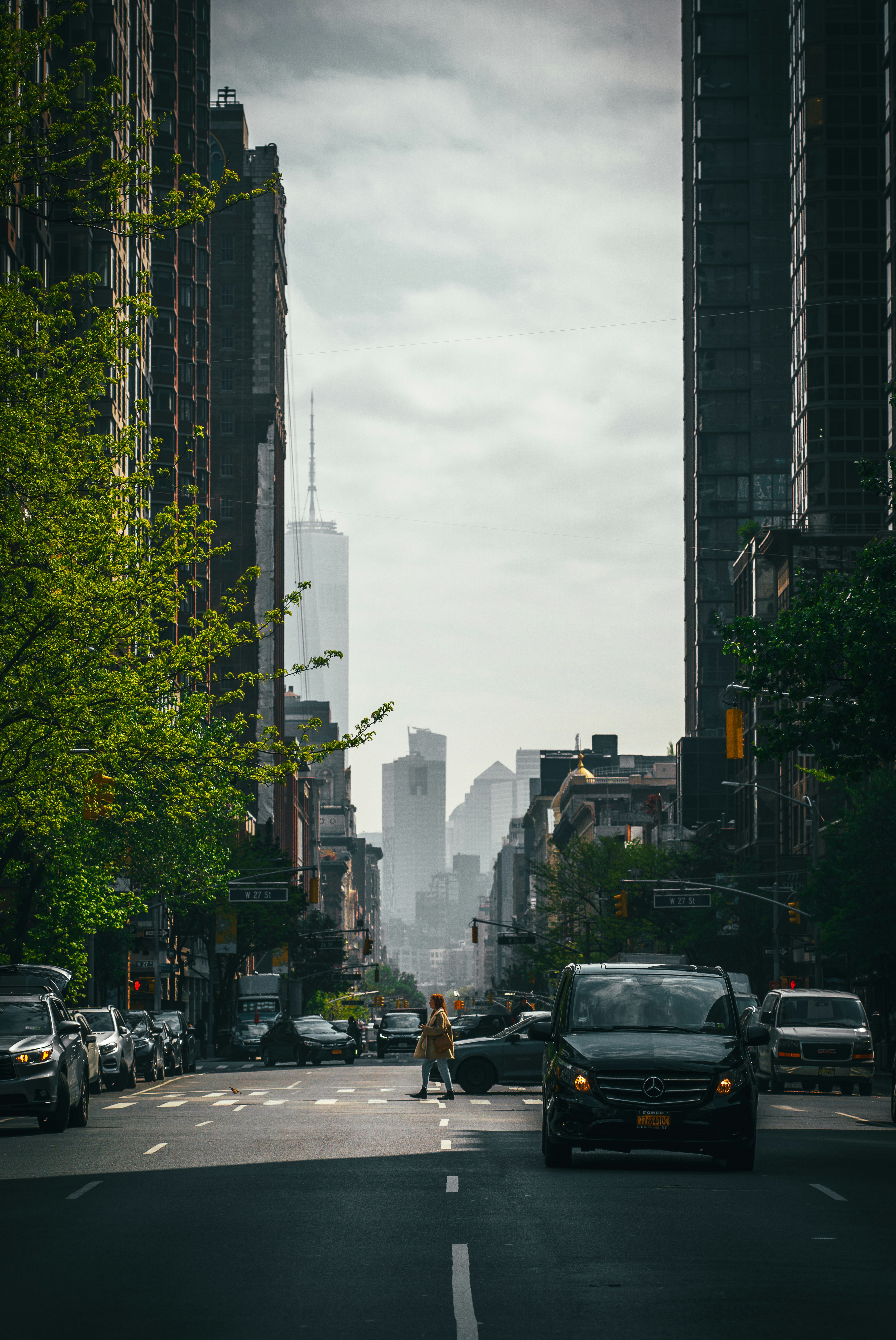 A lone figure crosses a bustling city street framed by towering buildings and lush greenery, with the skyline softly blurred in the background.