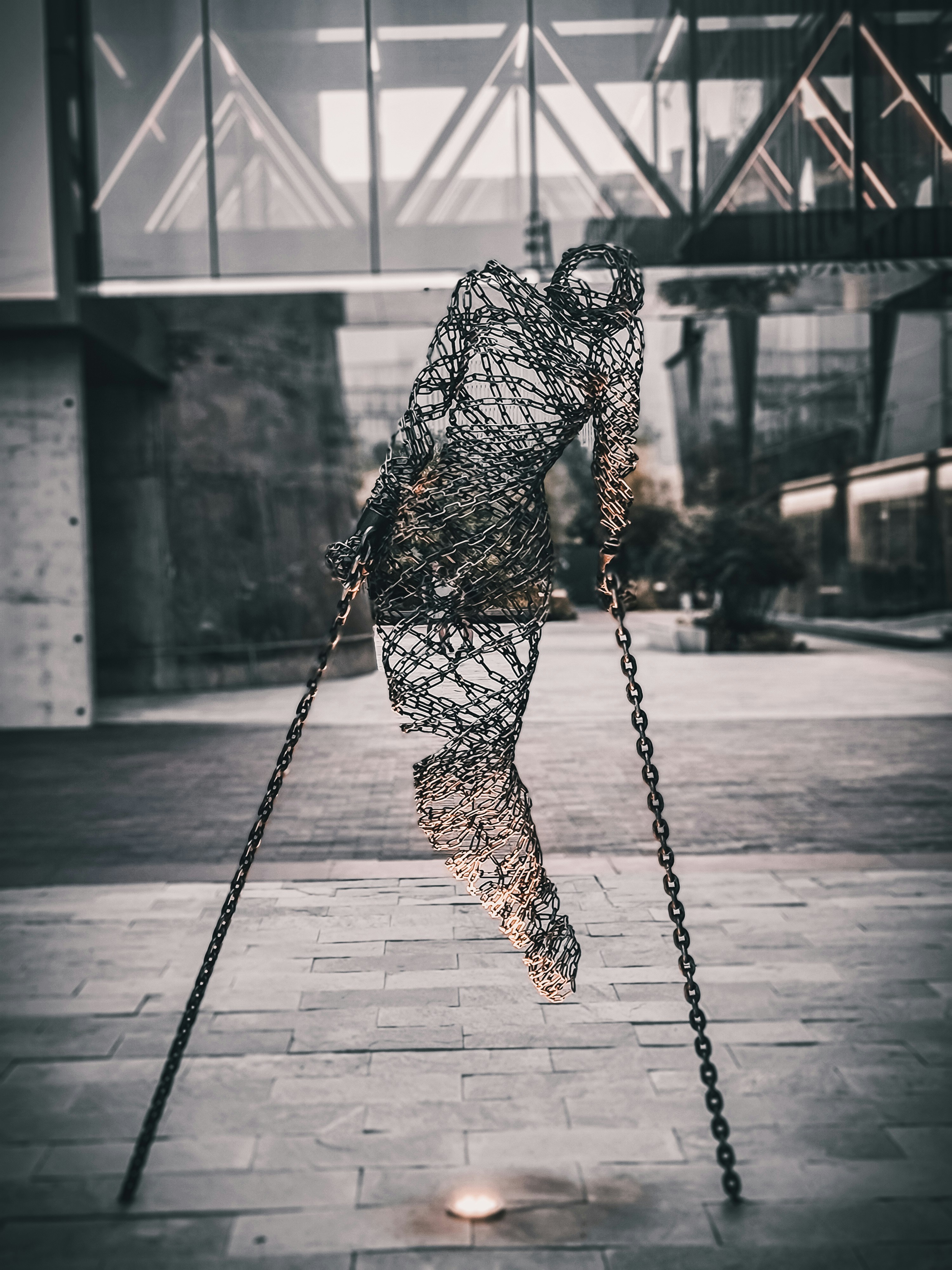 Weightless in Metal: A Wire-Mesh Figure Leaning Between Chains Under a Glass Atrium