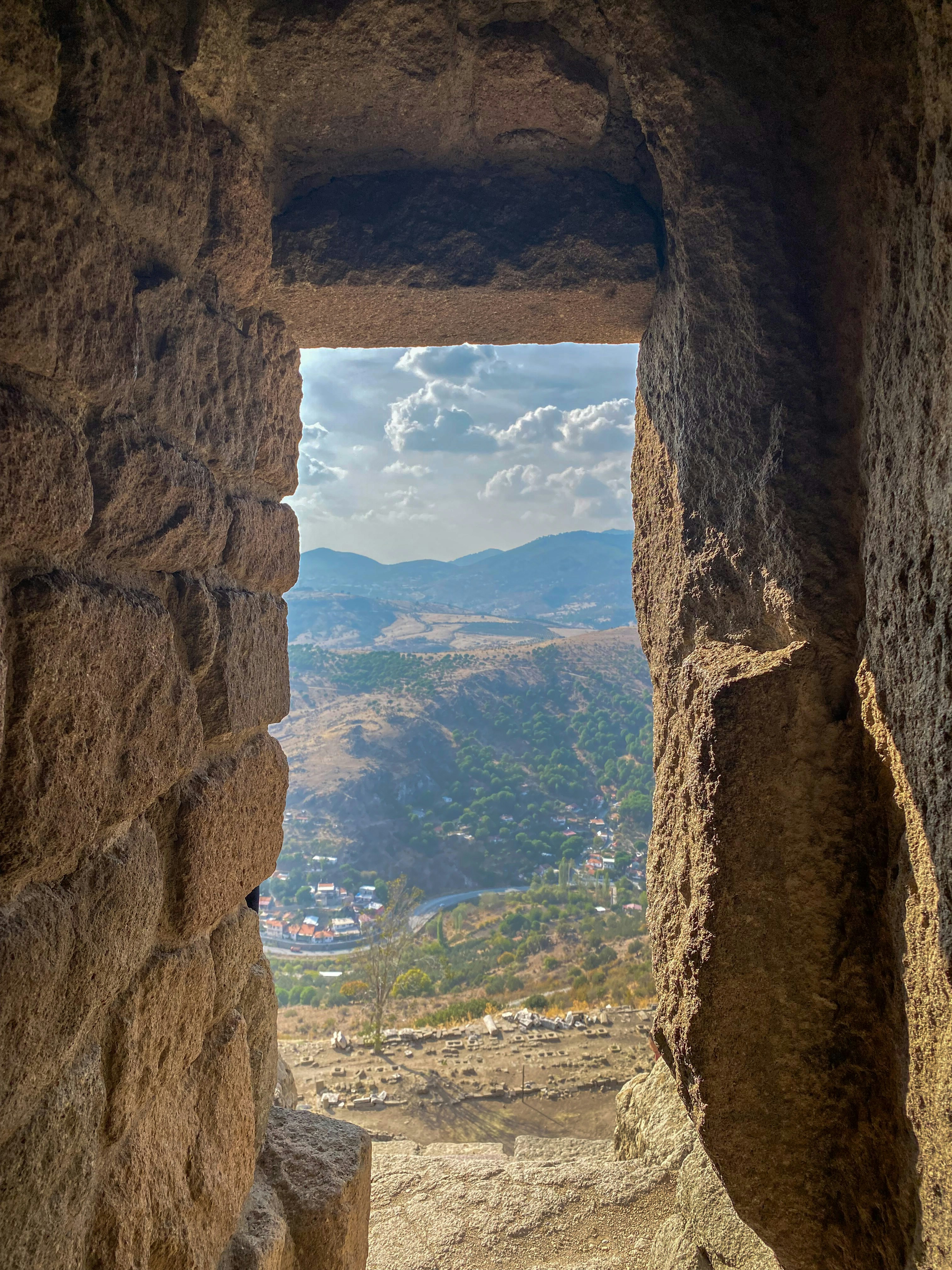 View from a stone window showcasing a valley with mountains and a small town below, illuminated by soft afternoon light.