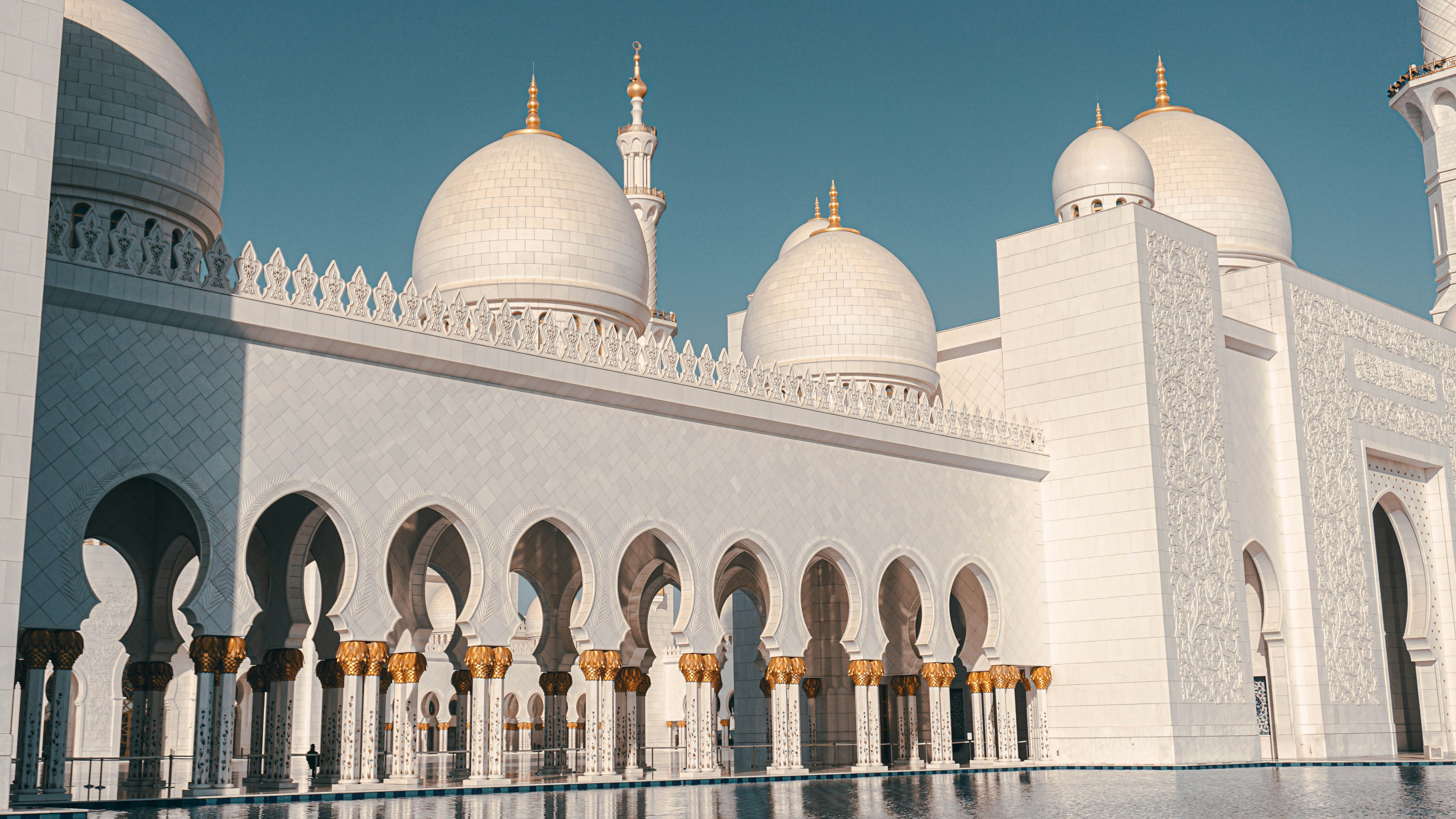 Intricate white domes and arches of a grand mosque reflecting in a tranquil pool under a clear blue sky.