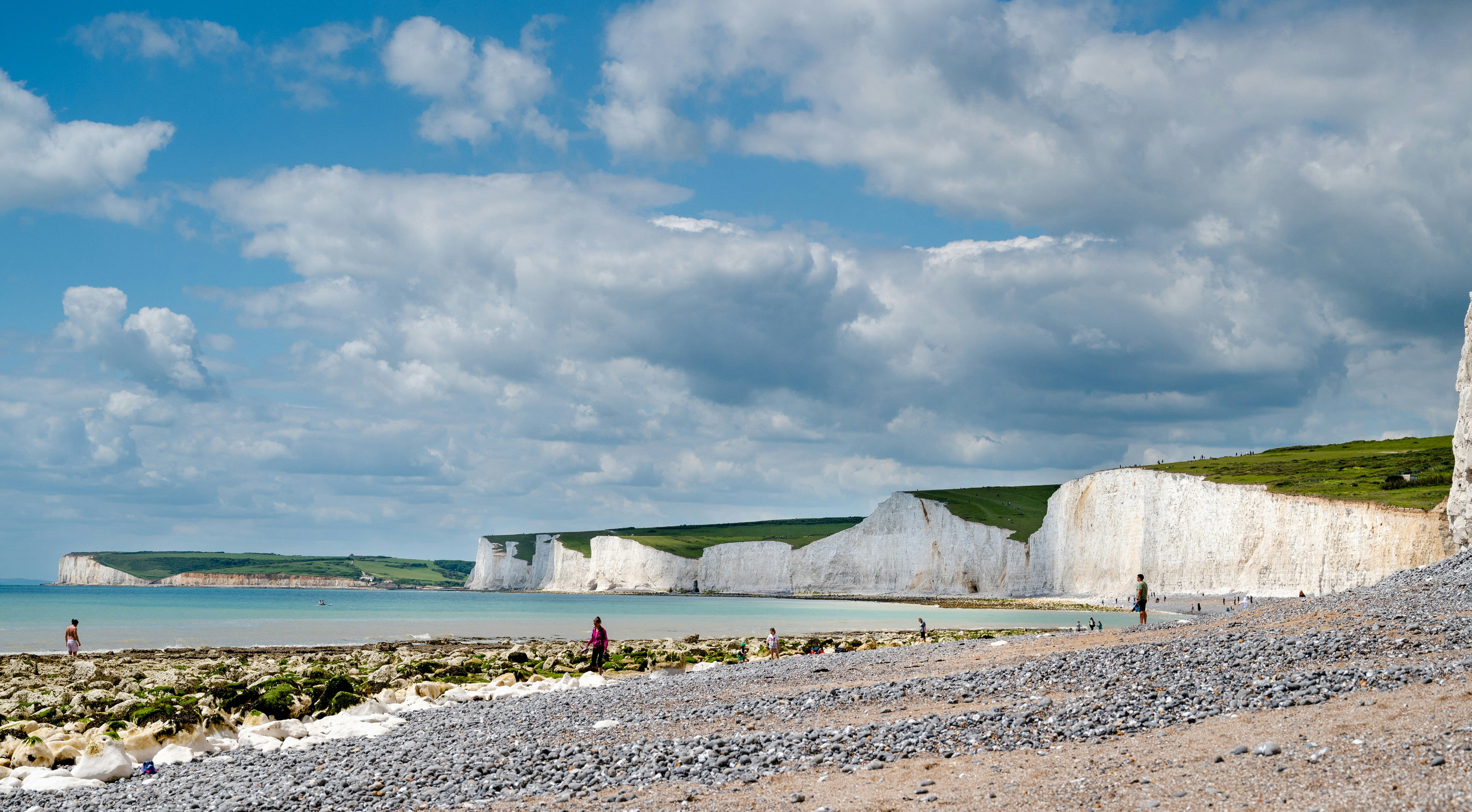 Expansive view of the White Cliffs along a pebbled beach under a dynamic sky, showcasing the interplay of land and sea. People stroll along the shore, emphasizing the scale of the cliffs.