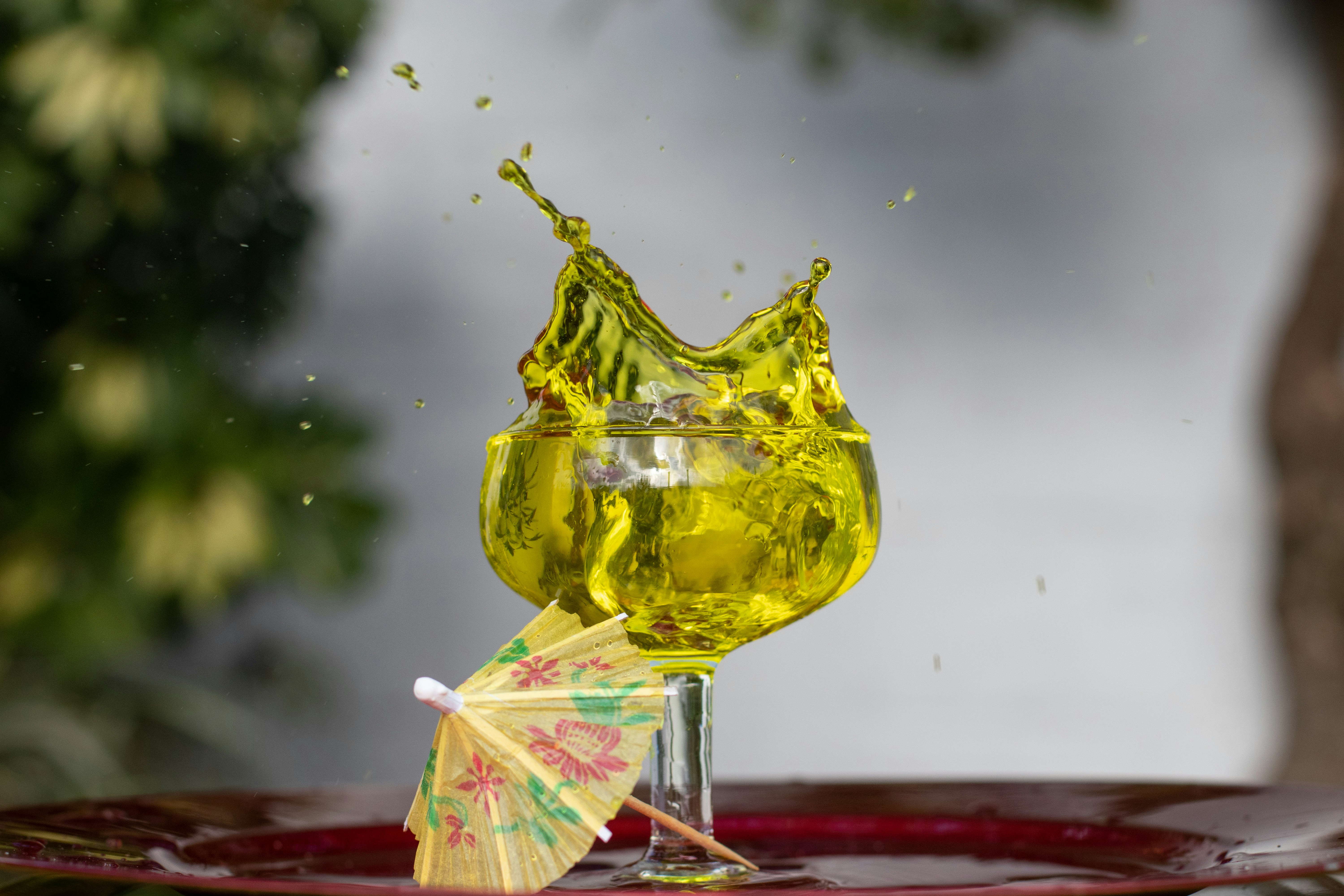 Vibrant yellow liquid splashes dramatically from a glass, accompanied by a colorful cocktail umbrella. The background features lush greenery, enhancing the tropical theme.
