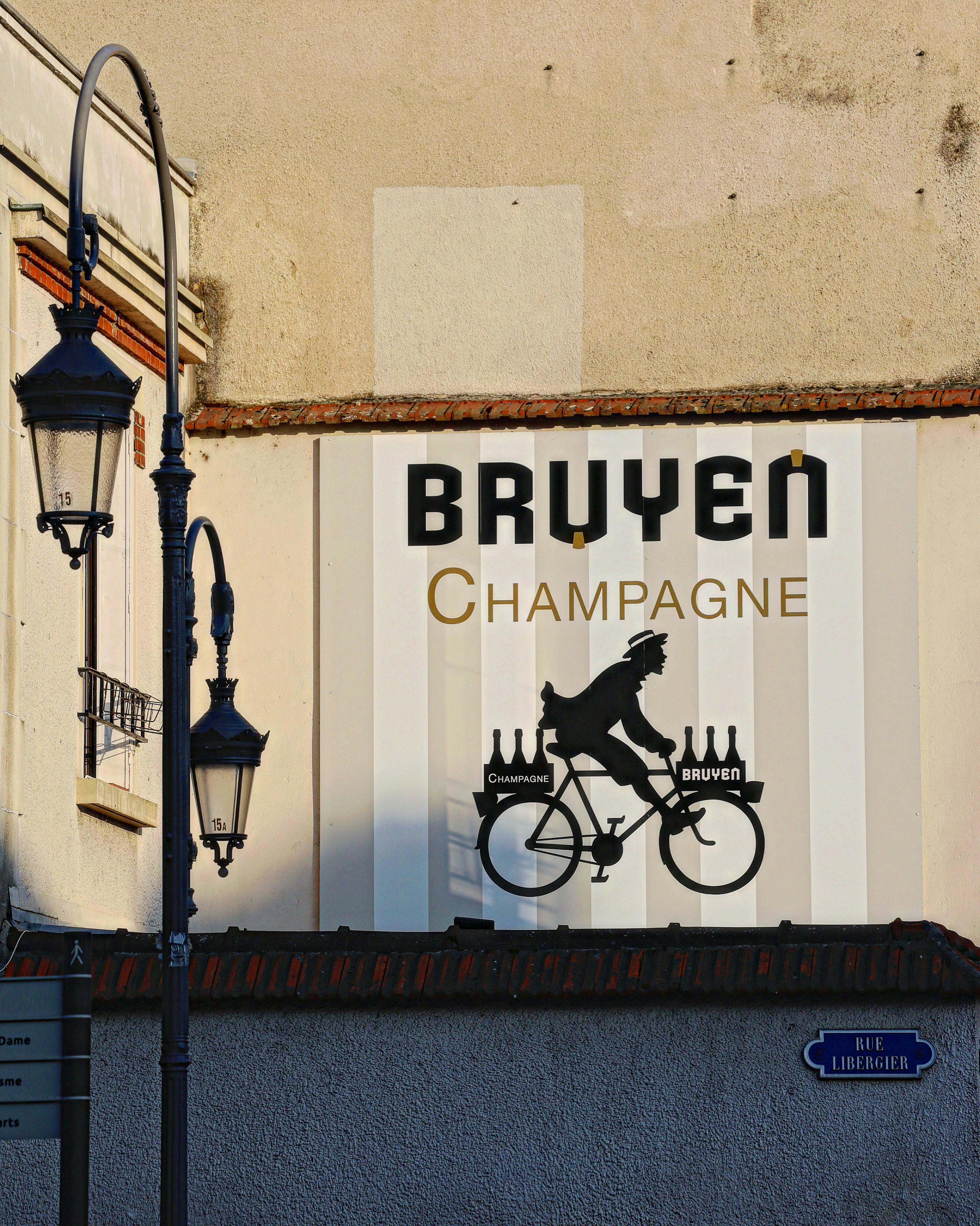 Wall mural of a cyclist carrying champagne bottles against a warm-toned building facade.
