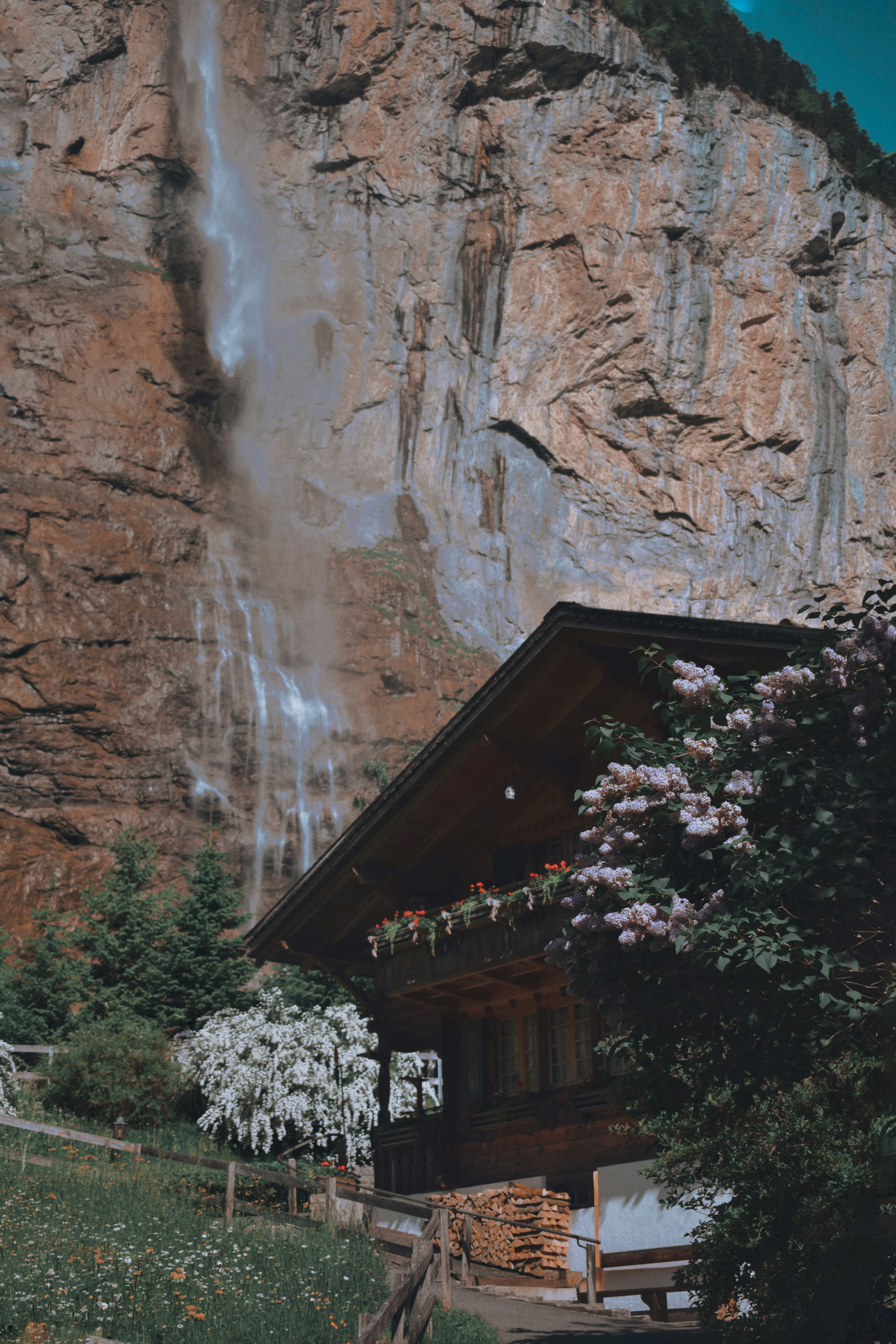 Charming wooden chalet nestled against a towering cliff, adorned with vibrant flowers and a cascading waterfall in the background.