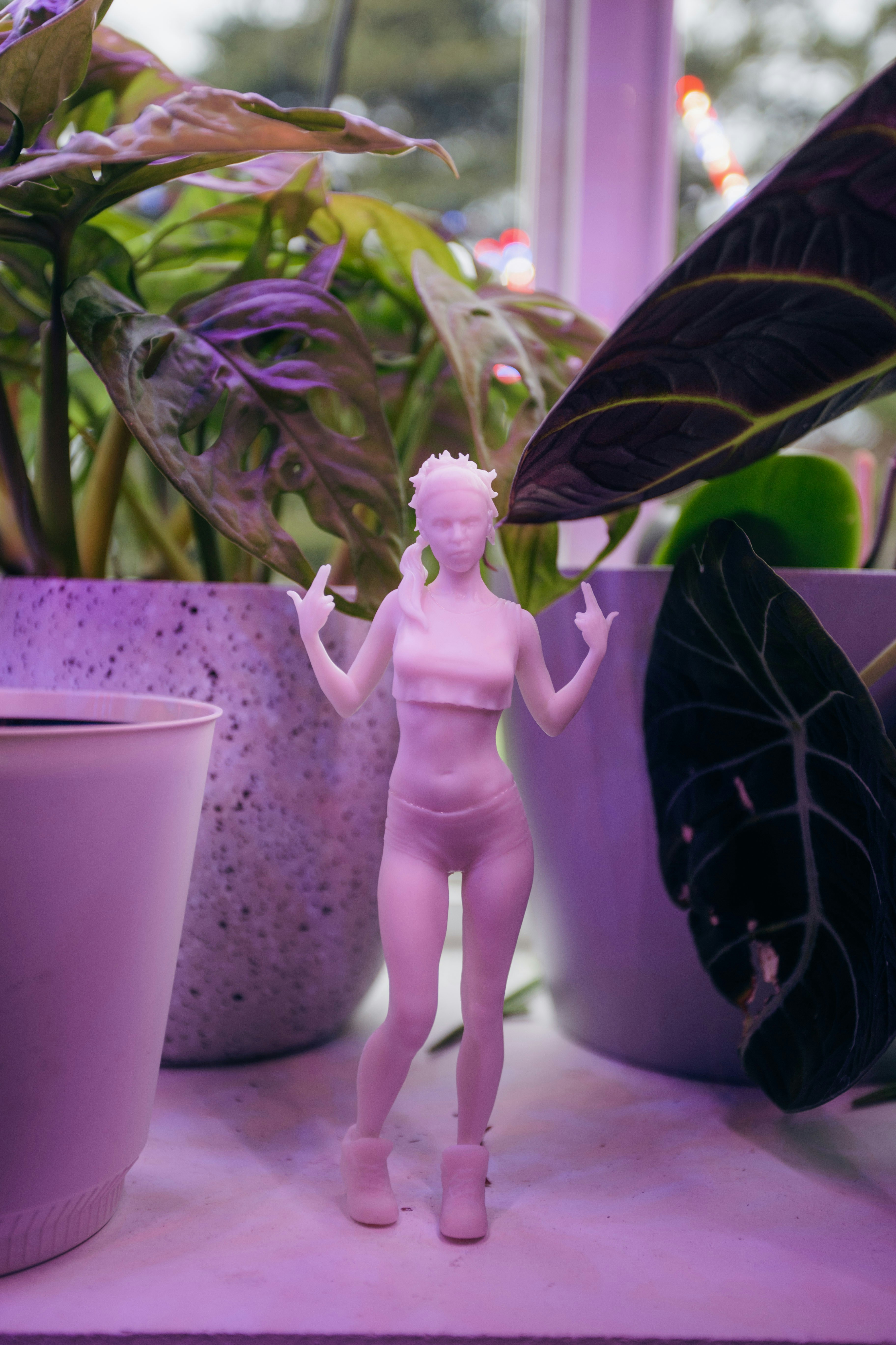 A stylized figurine stands amidst lush plants, exuding a playful attitude with a bold gesture. The vibrant greens and soft lighting create a lively atmosphere.
