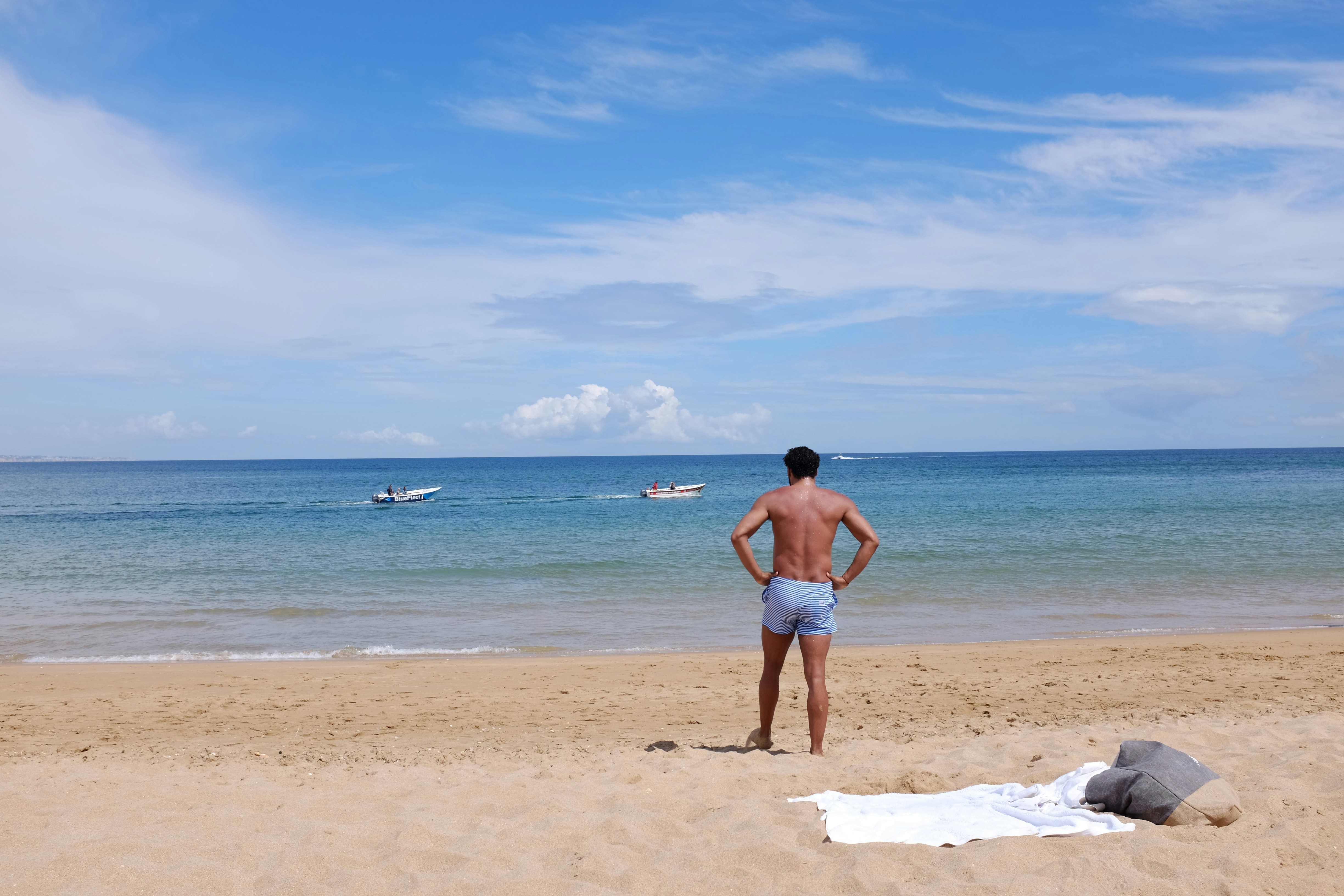 A man in blue swim trunks stands on a Portugal beach, looking at the ocean.