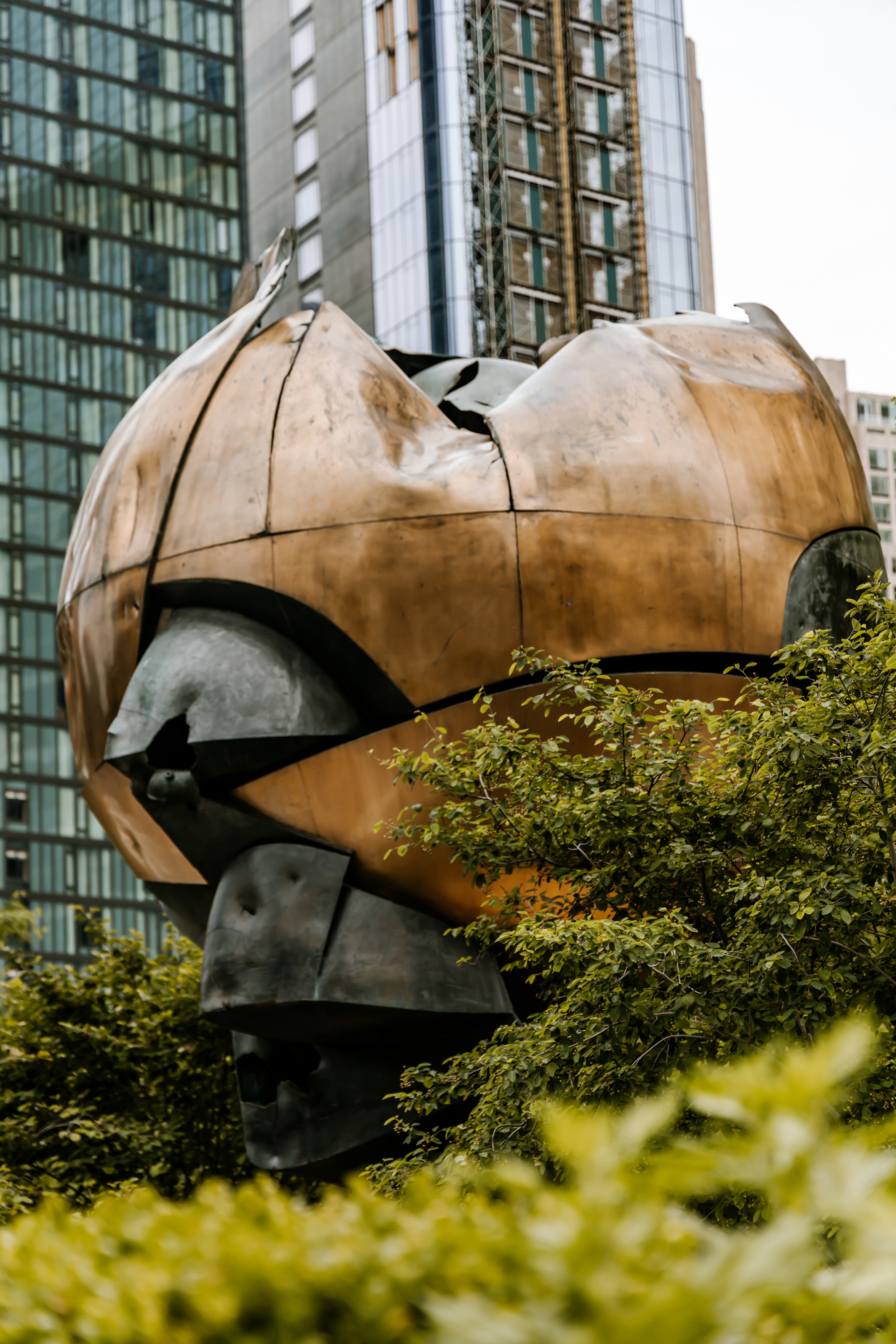 A striking sculpture resembling a fragmented sphere, partially obscured by lush greenery, juxtaposed against modern skyscrapers.