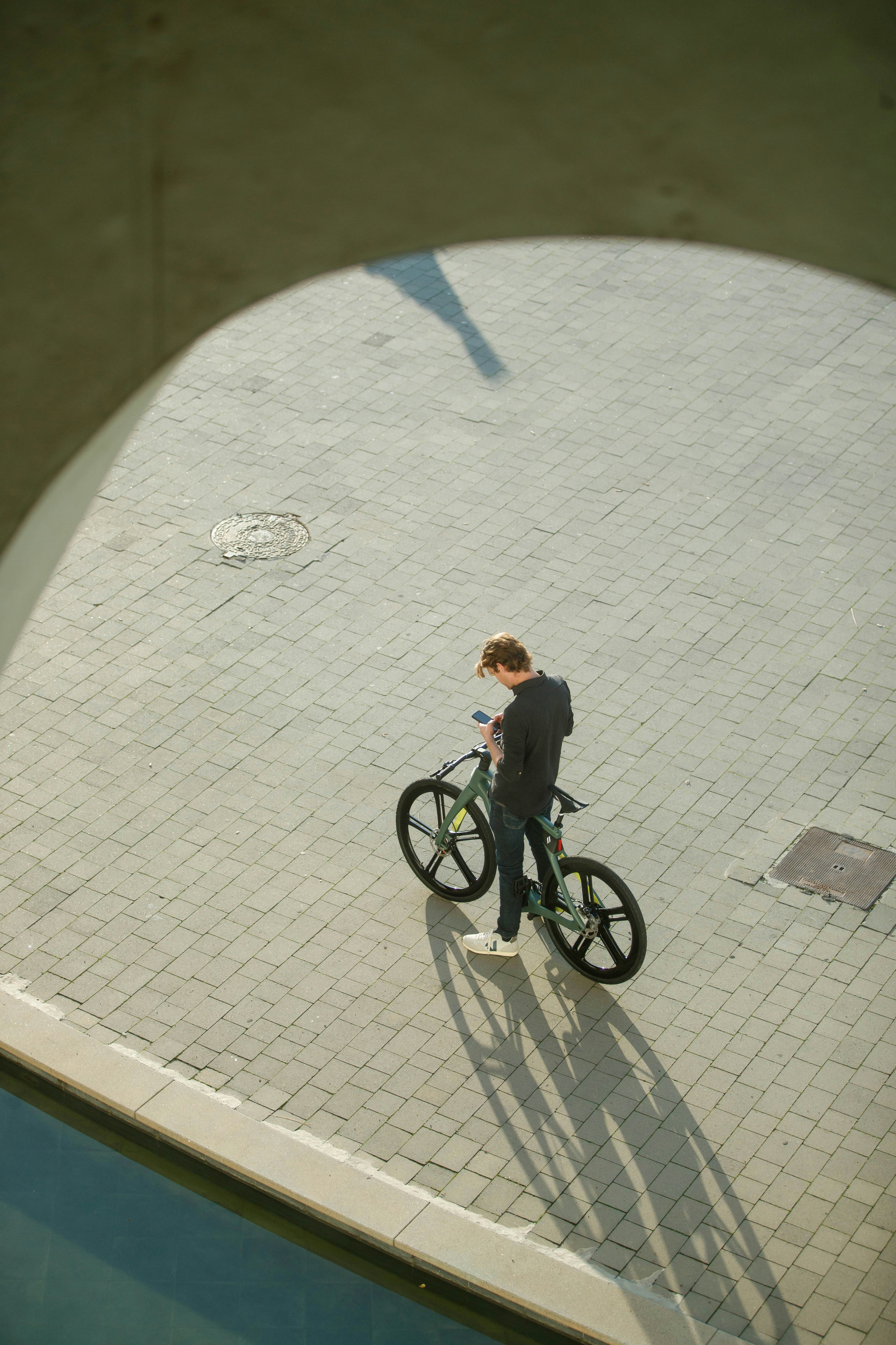Young man checking his phone while standing beside a bicycle on a cobblestone street, framed by an architectural arch. 