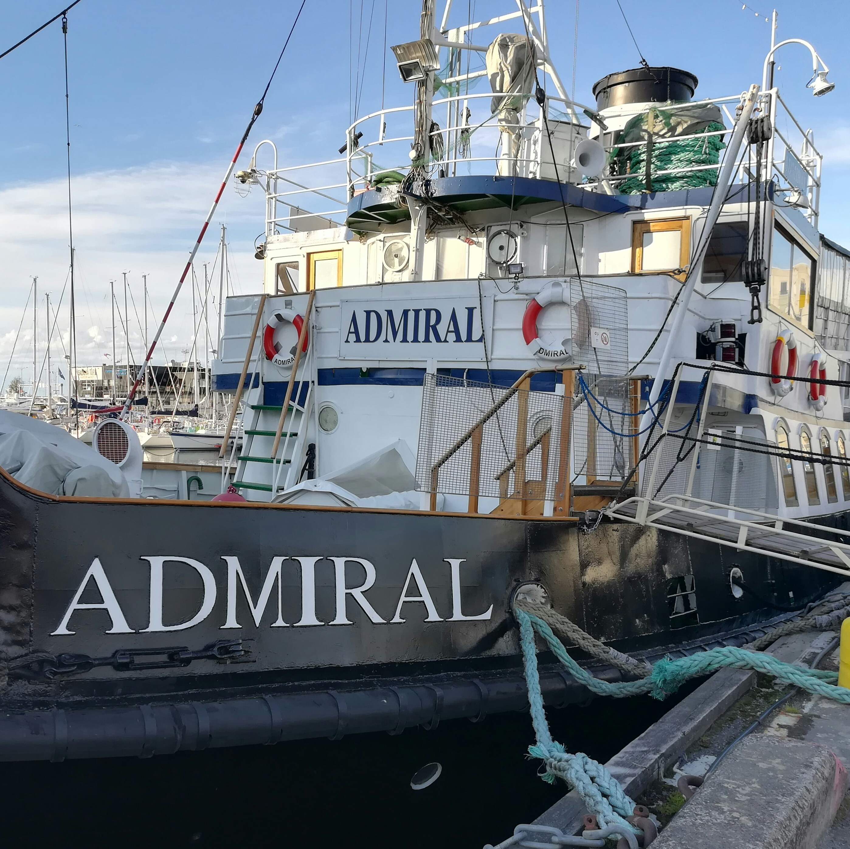 A docked tugboat named Admiral dominates the scene, with bold lettering on the hull and a clear gangway leading up to a multi-level wheelhouse against a bright sky. This is a maritime vessel photograph.