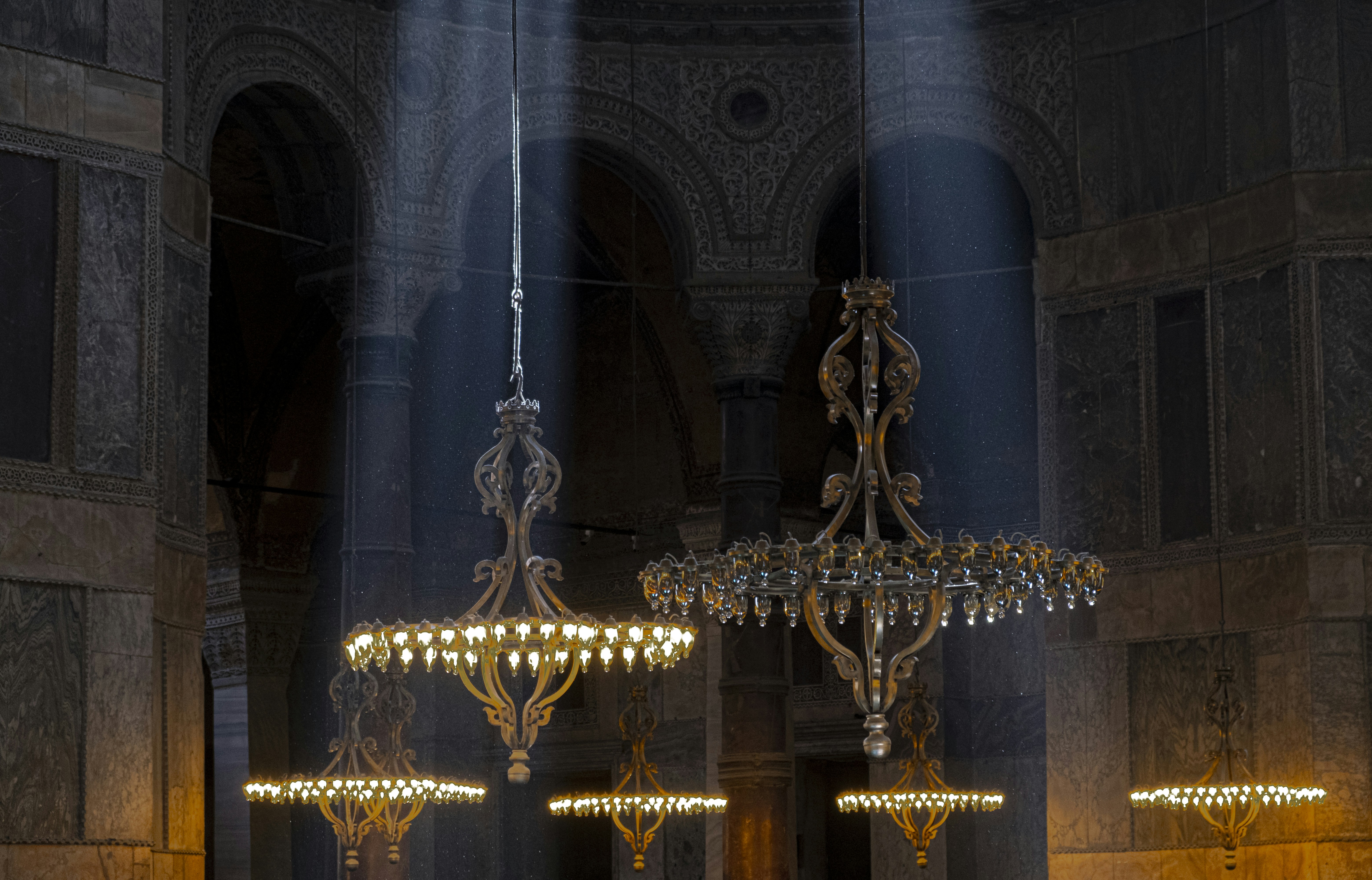 Intricate chandeliers suspended in a grand hall, illuminated by beams of light filtering through the architecture. The ornate details and soft glow create a serene atmosphere.