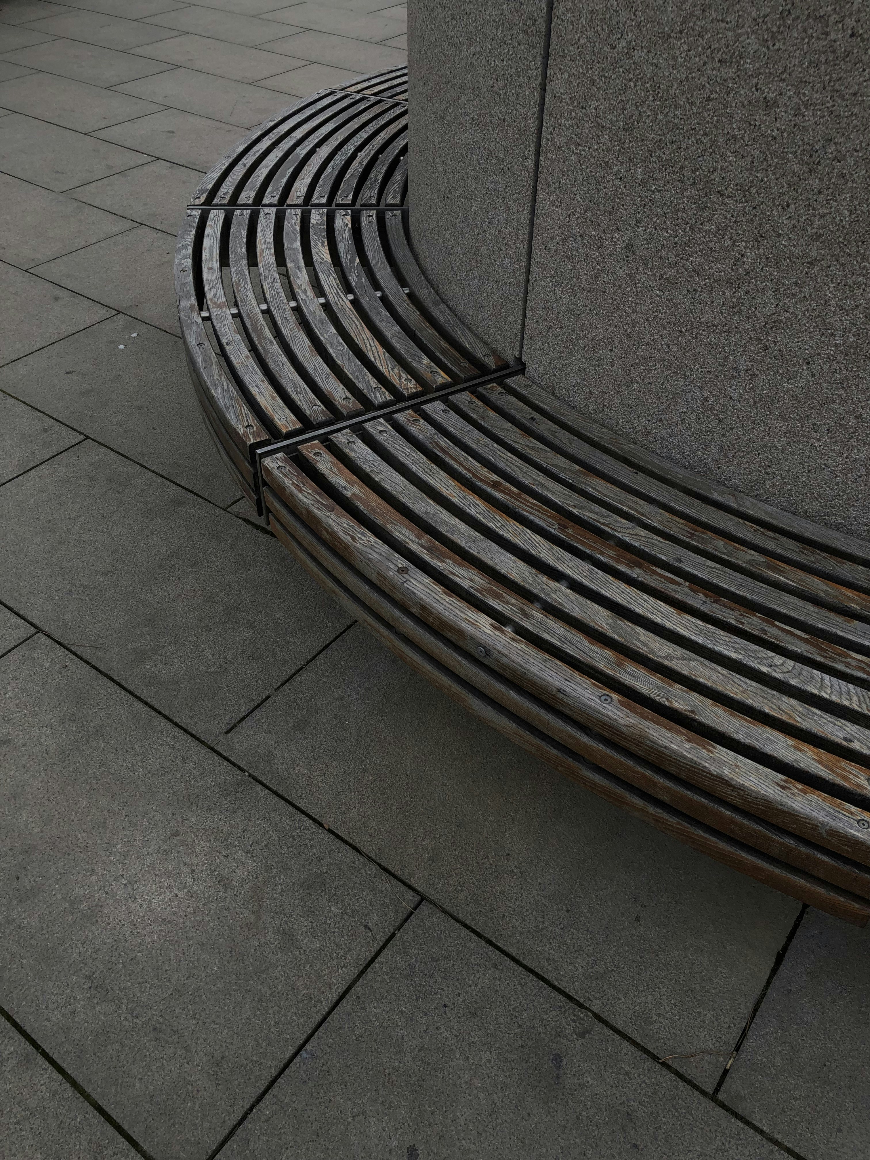 Wooden slatted bench curving around a stone pillar on a paved surface.