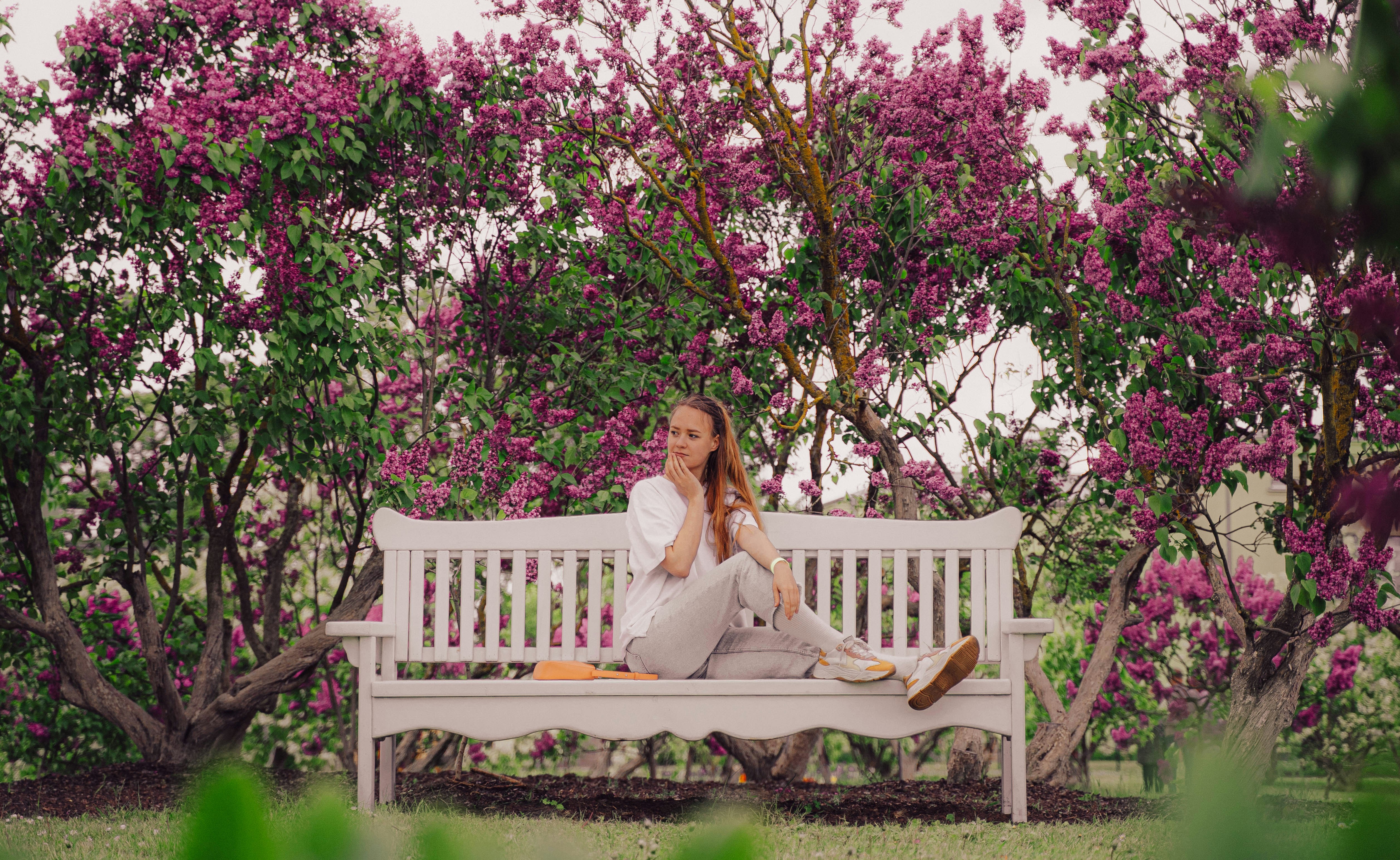 A woman in casual attire sits pensively on a white bench surrounded by vibrant lilac blooms, creating a tranquil atmosphere.