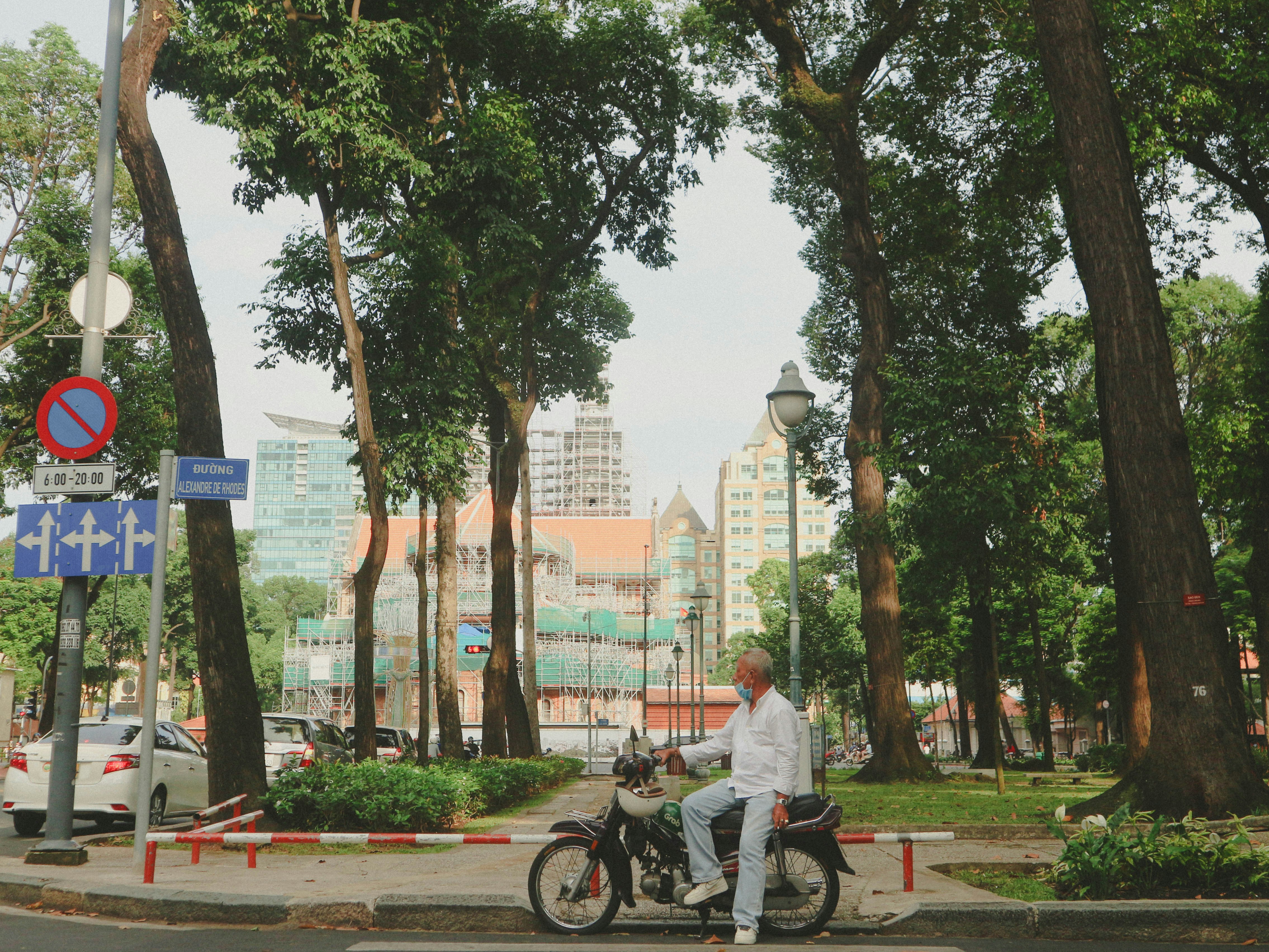 An elderly man in a white outfit sits on a motorcycle amidst a lush park setting, framed by towering trees and urban buildings in the background.