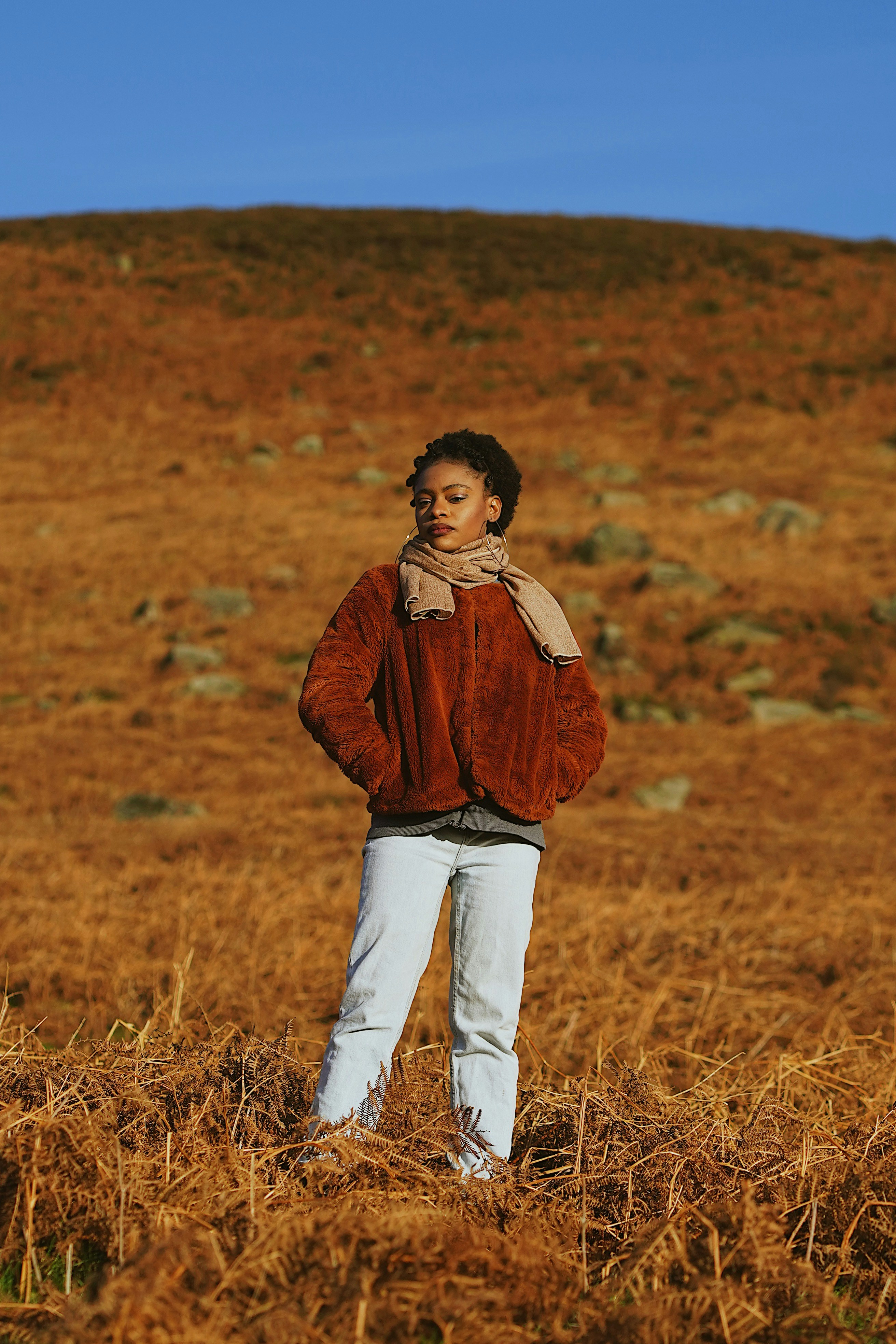 Individual standing confidently in a sunlit field of golden ferns, wearing a cozy brown jacket and scarf. Natural landscape features rolling hills in the background.