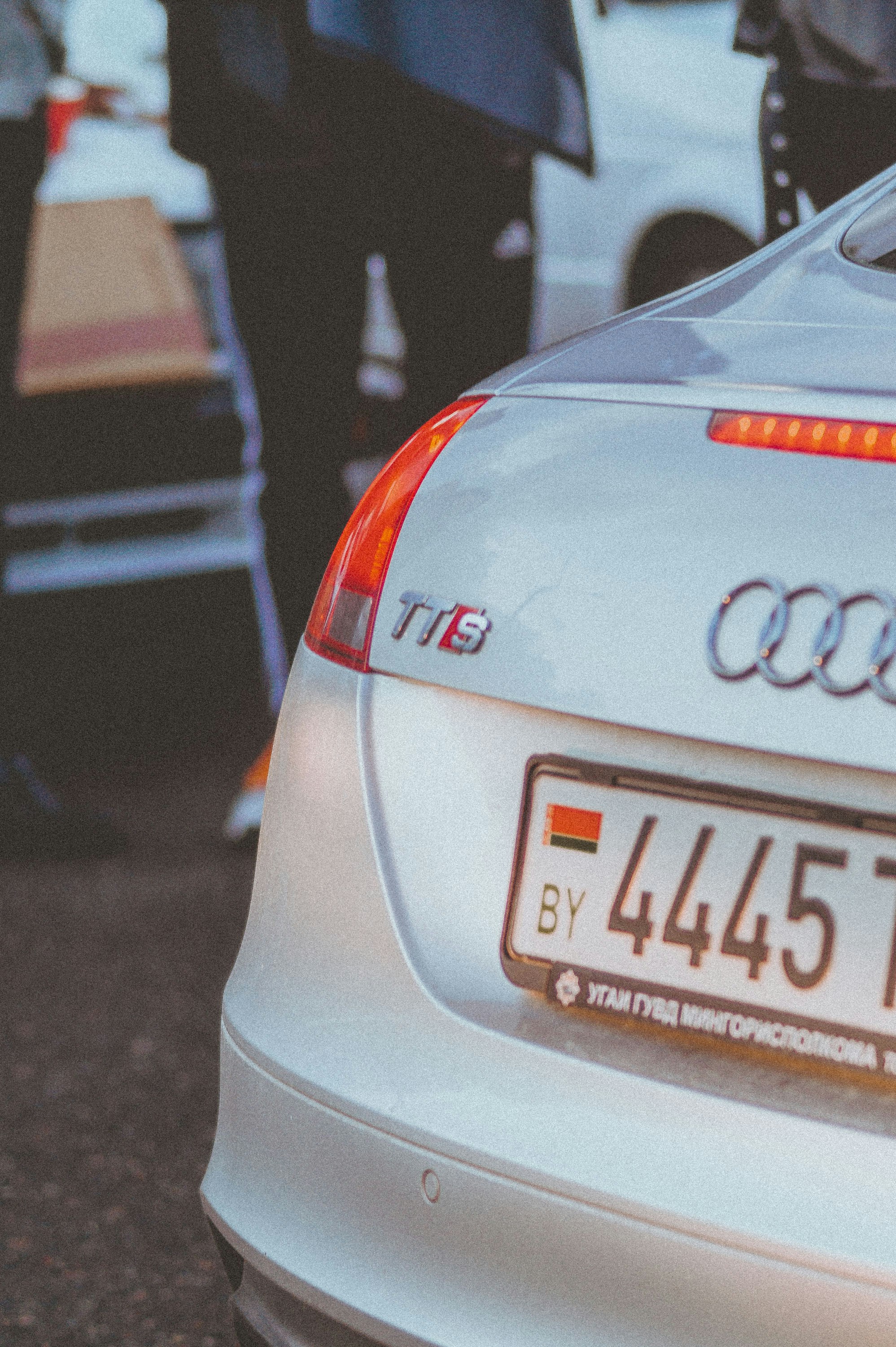 Curved taillight and license plate narrative on a silver Audi TT-S in a bustling parking scene