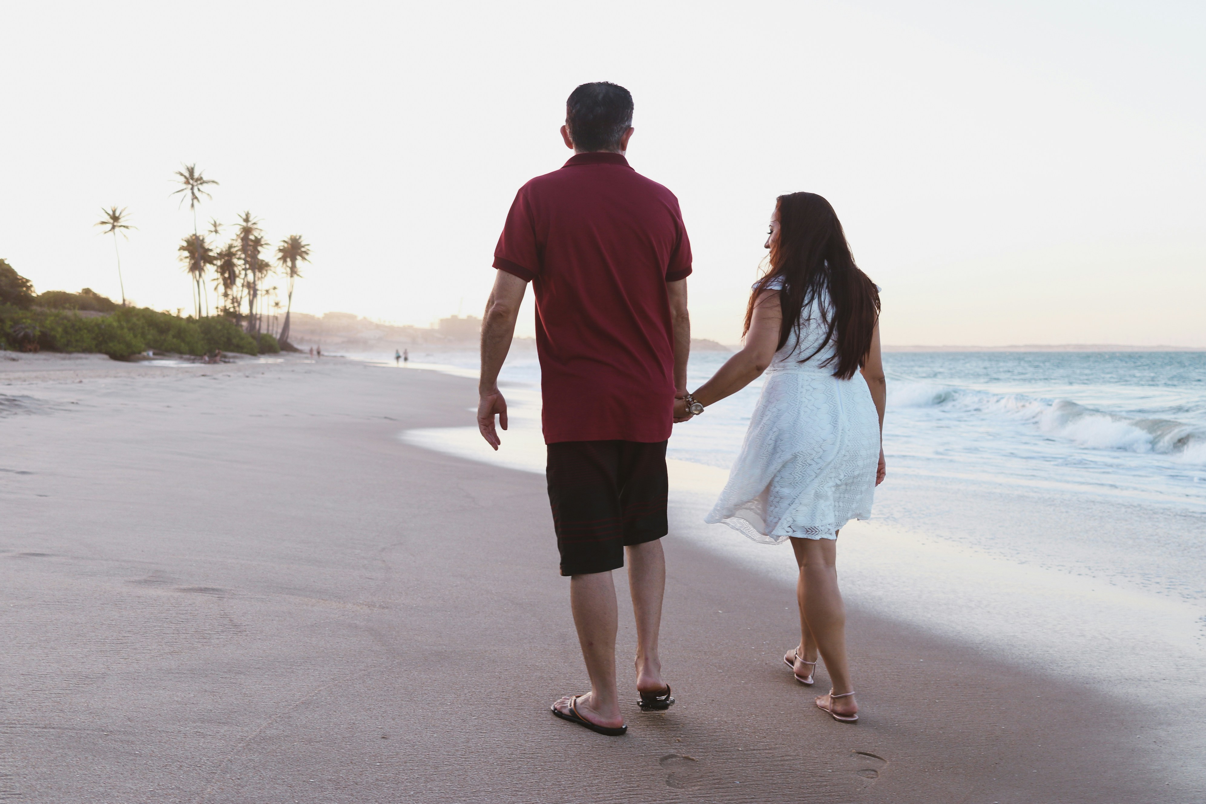 Couple walking hand in hand along a sandy beach with gentle waves and palm trees in the distance.