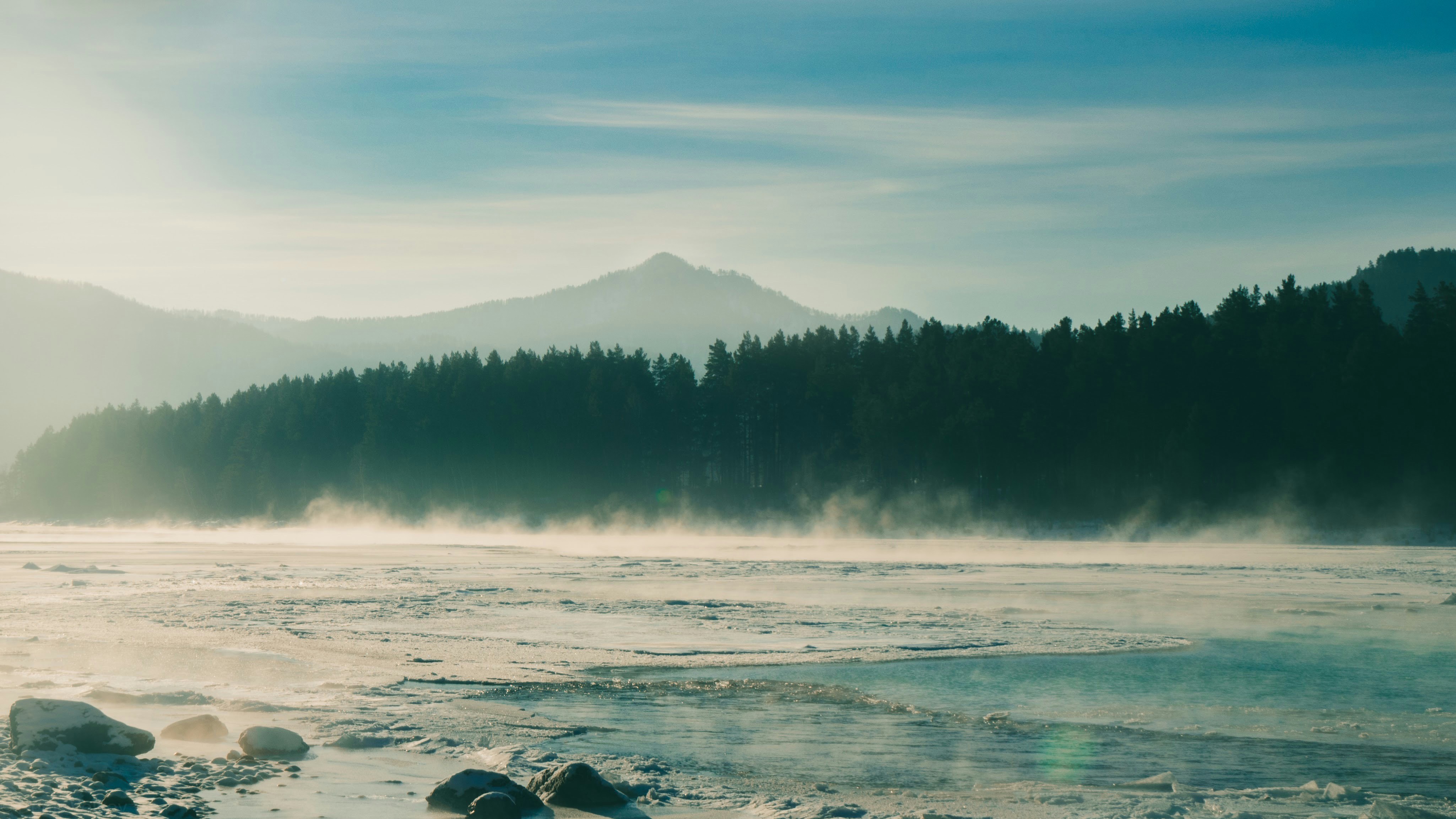 Fog drifts over a tranquil lake with distant mountains under a blue sky.