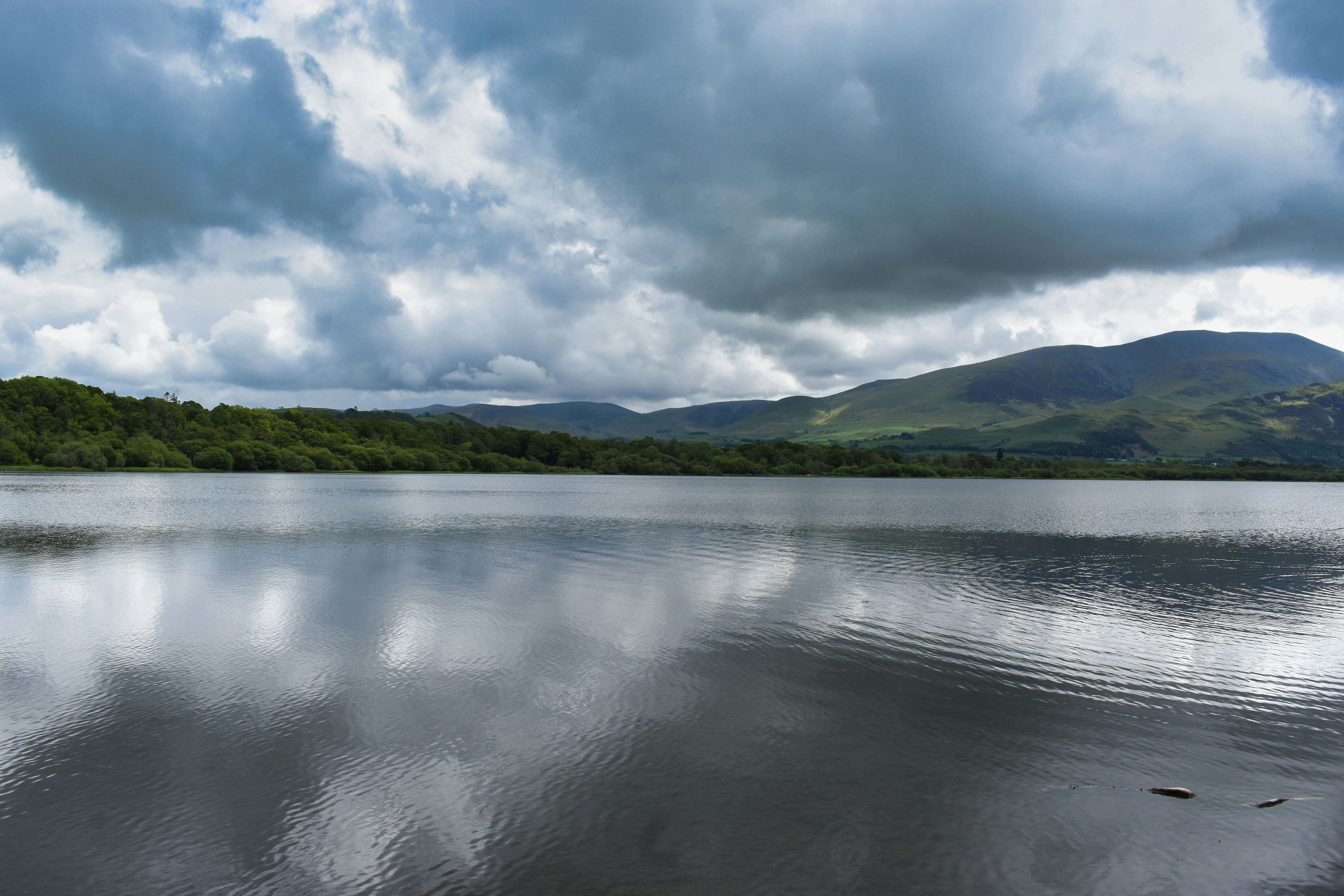 Tranquil lake reflecting dramatic clouds and distant hills, surrounded by lush greenery.