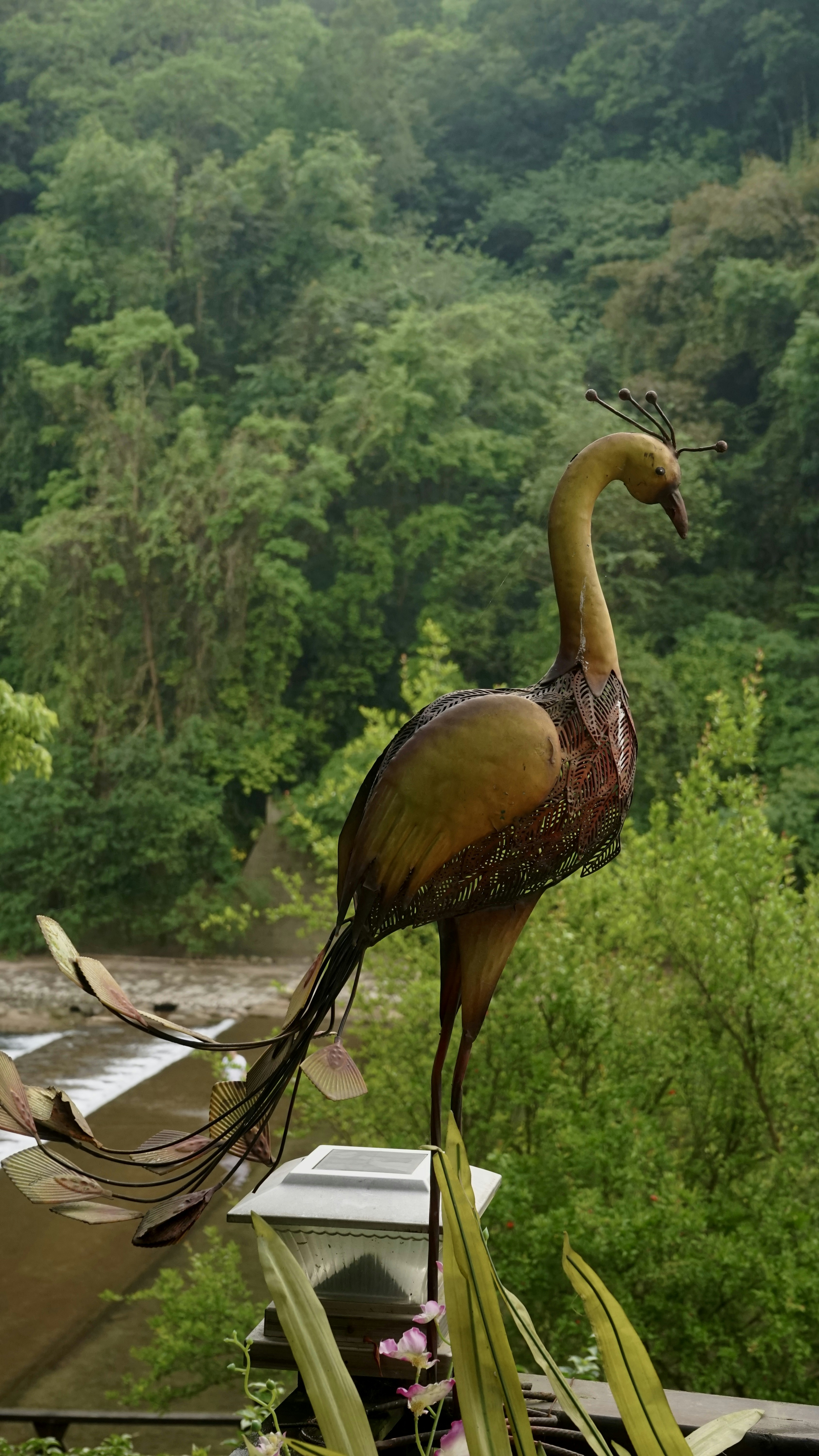 A beautifully crafted metal sculpture of a bird stands proudly among greenery, overlooking a serene riverbank.