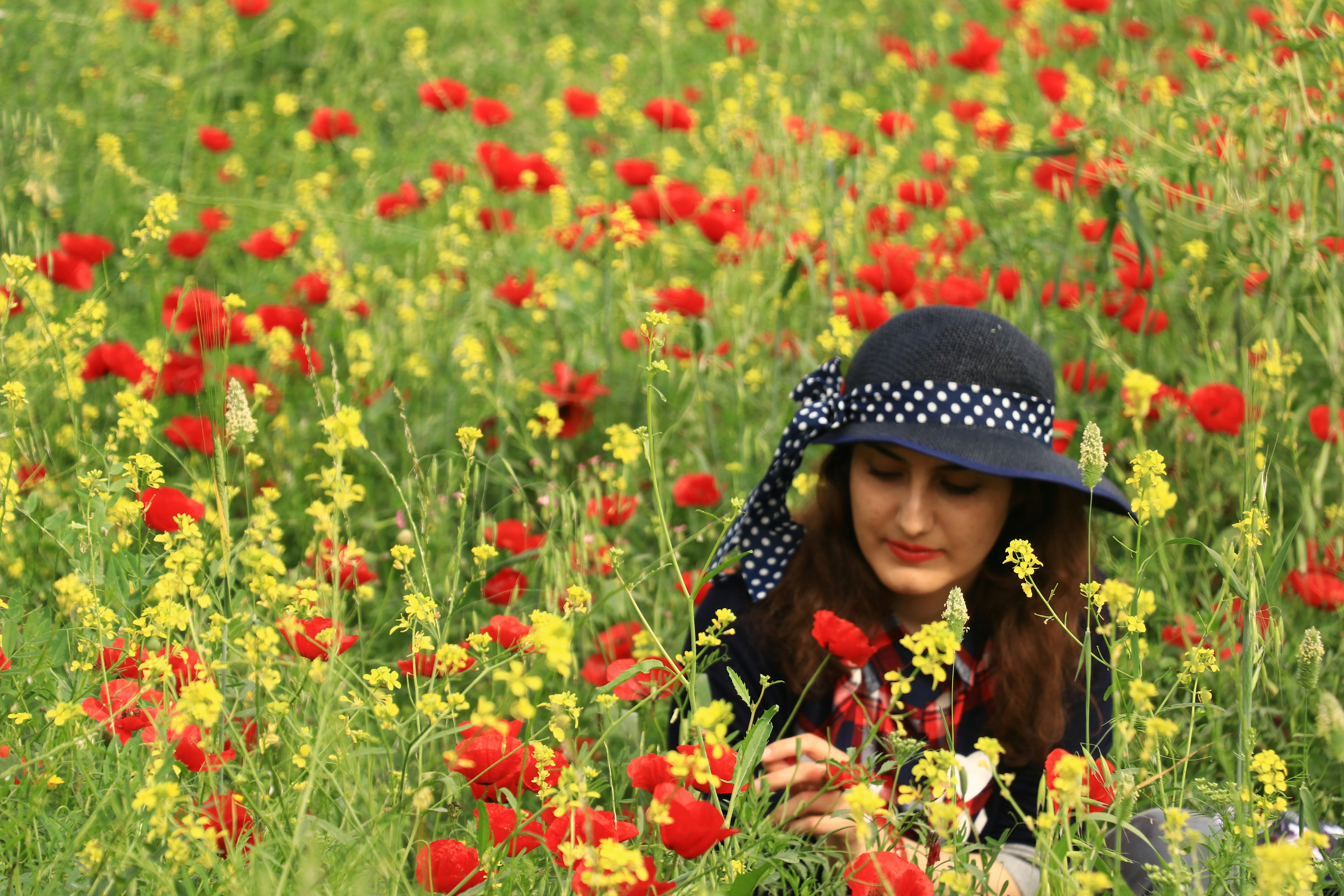 A woman in a black hat adorned with a polka dot ribbon kneels amidst vibrant red poppies and yellow wildflowers, immersed in nature's beauty.