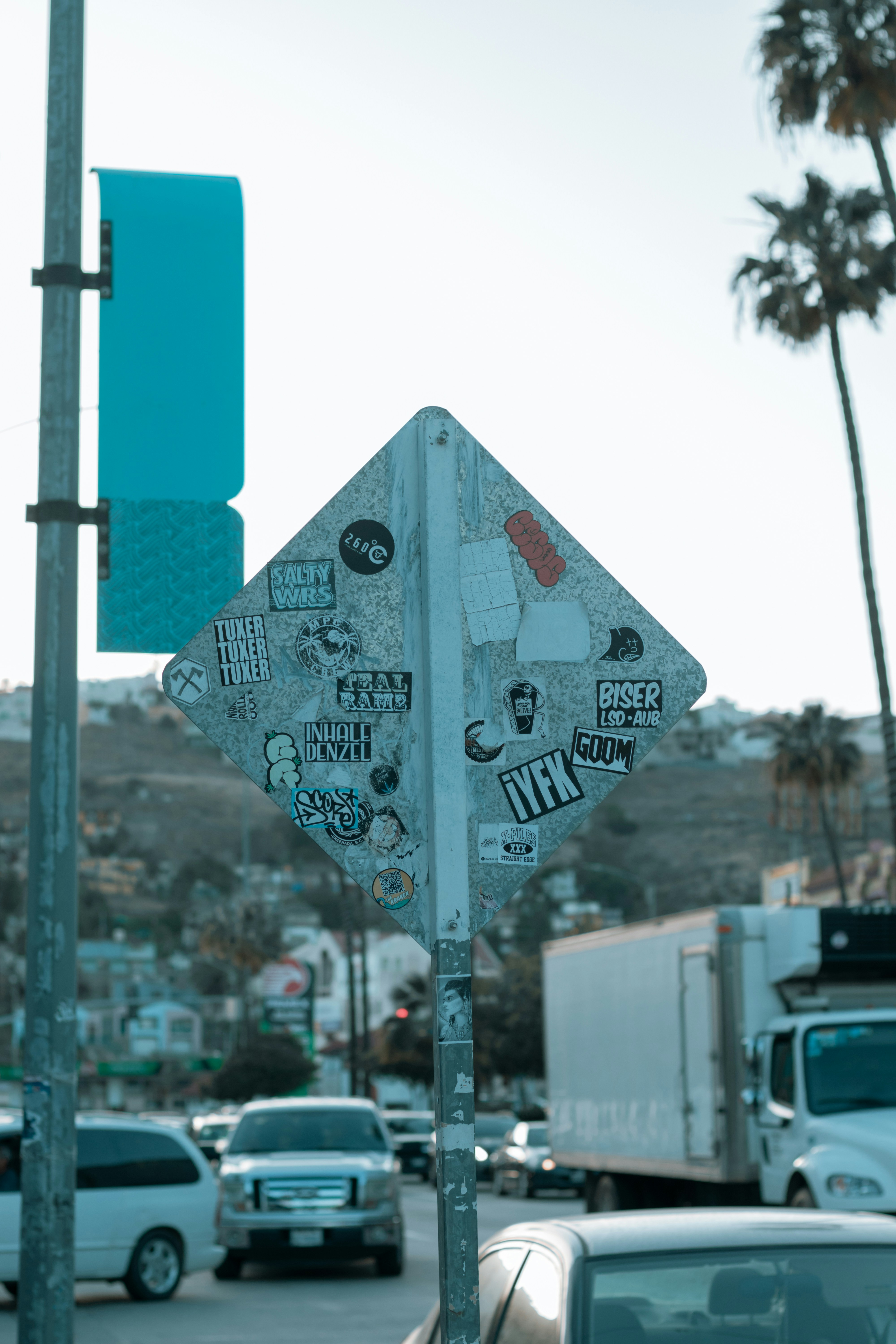 Stickered Diamond Sign: Weathered Beacon Amid Palm-Lined Traffic