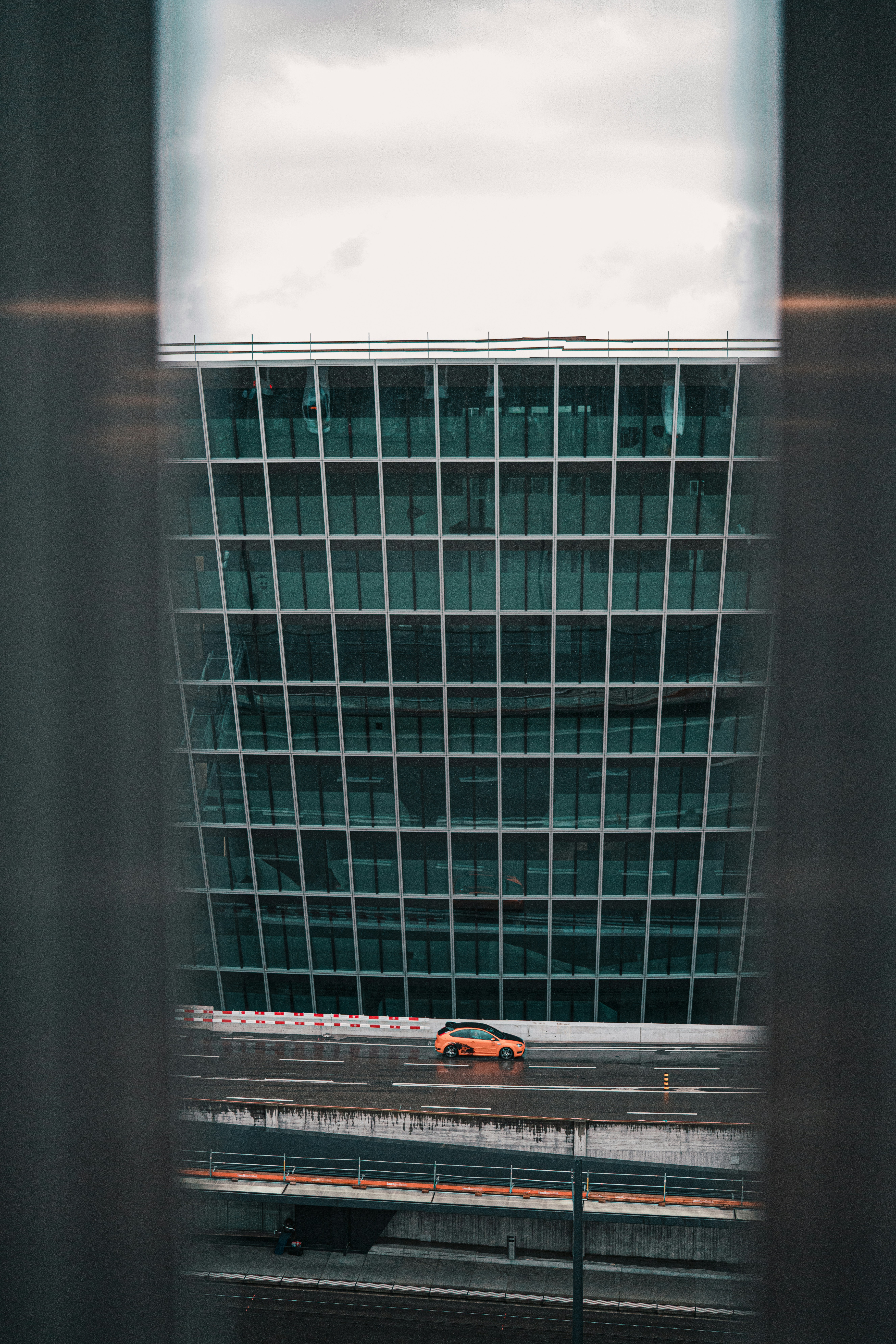 A vibrant orange car navigates a sleek, modern roadway beside a glass-paneled building, framed by vertical metal structures.