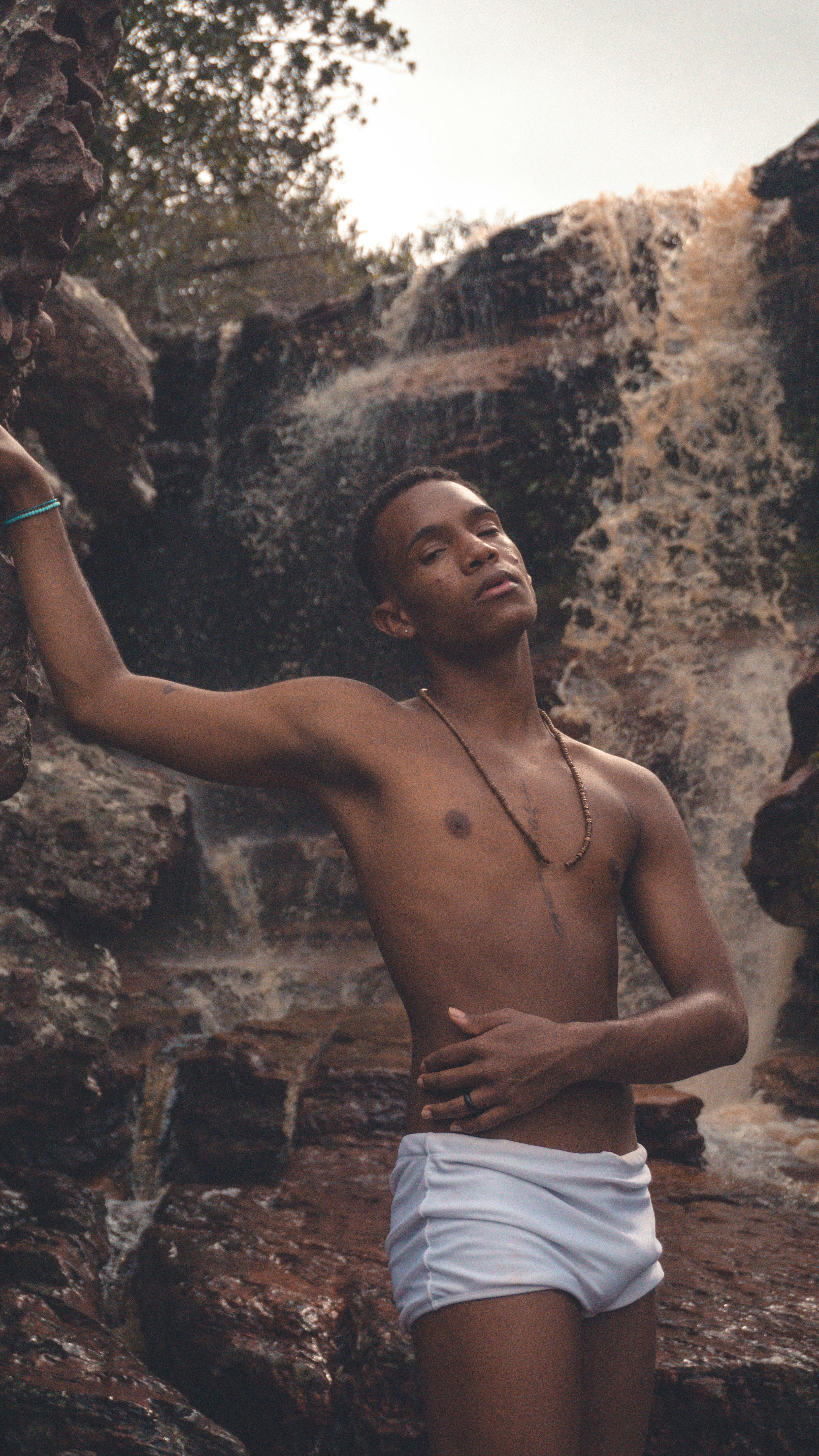 Young man posing confidently by a waterfall, showcasing the harmony between human form and natural beauty.