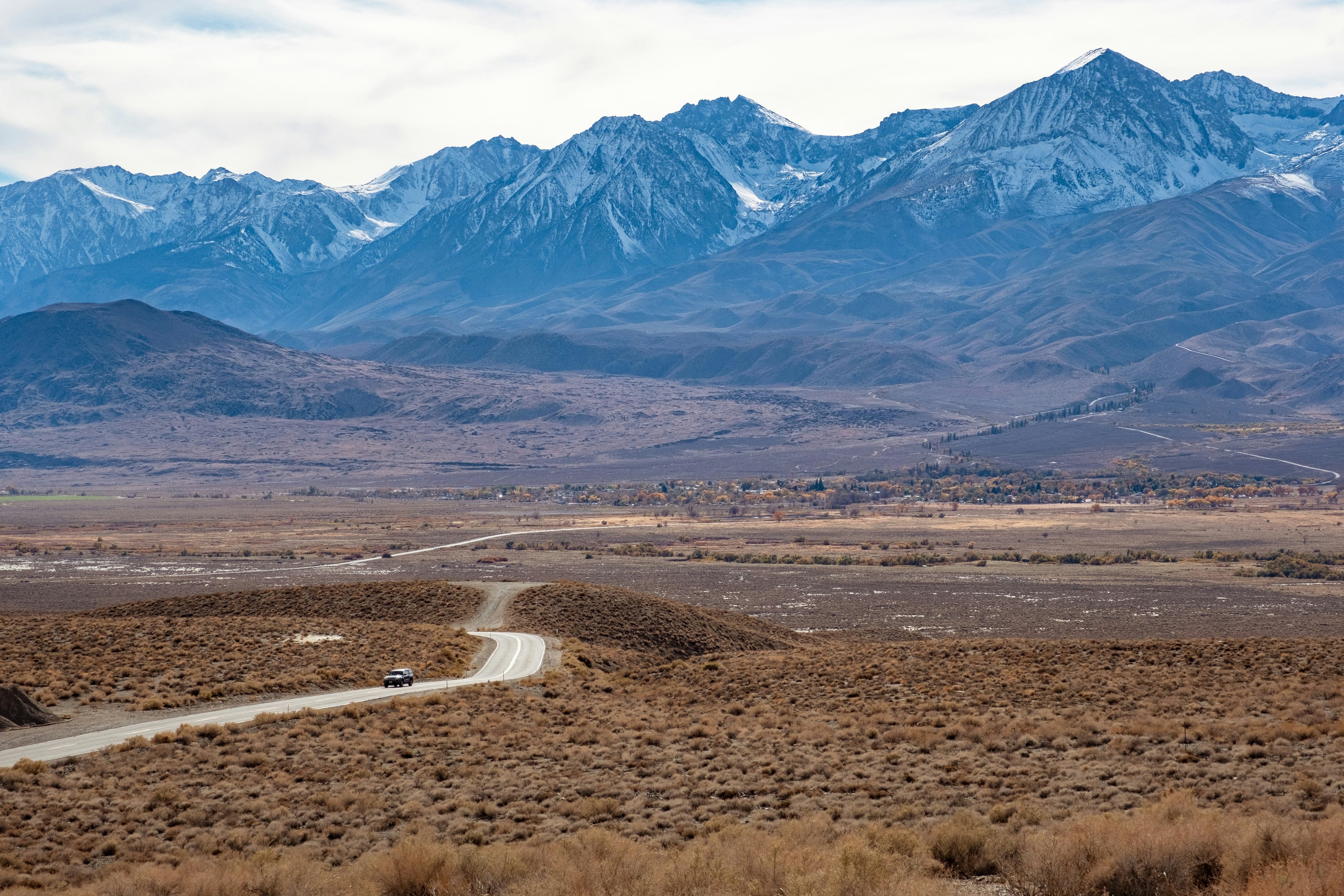 A solitary vehicle navigates a winding road amidst expansive desert terrain and towering snow-capped mountains in the background.