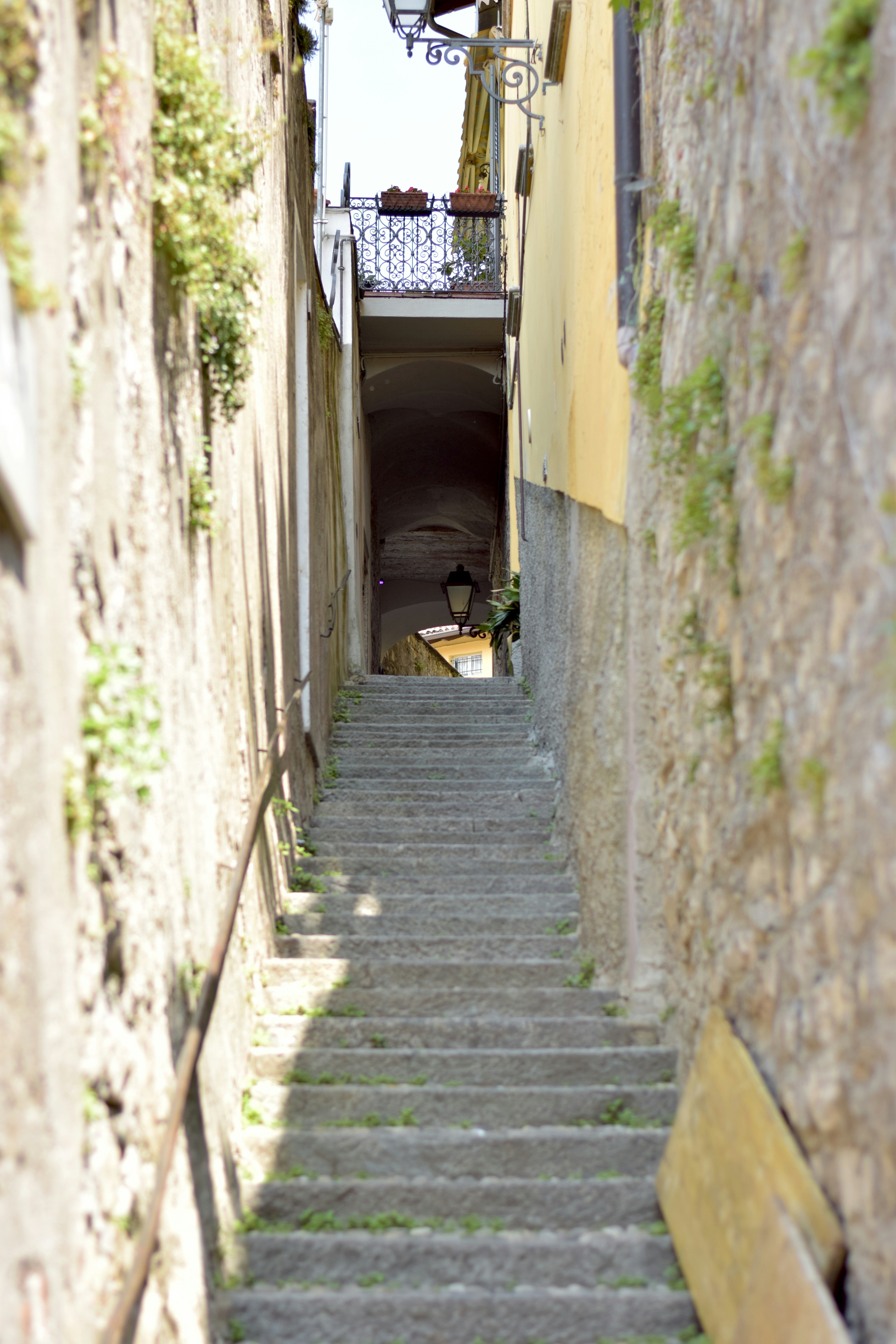 Narrow stone staircase leading up through a quaint alley, framed by weathered walls and greenery, inviting exploration. A lantern hangs above, hinting at the charm above.