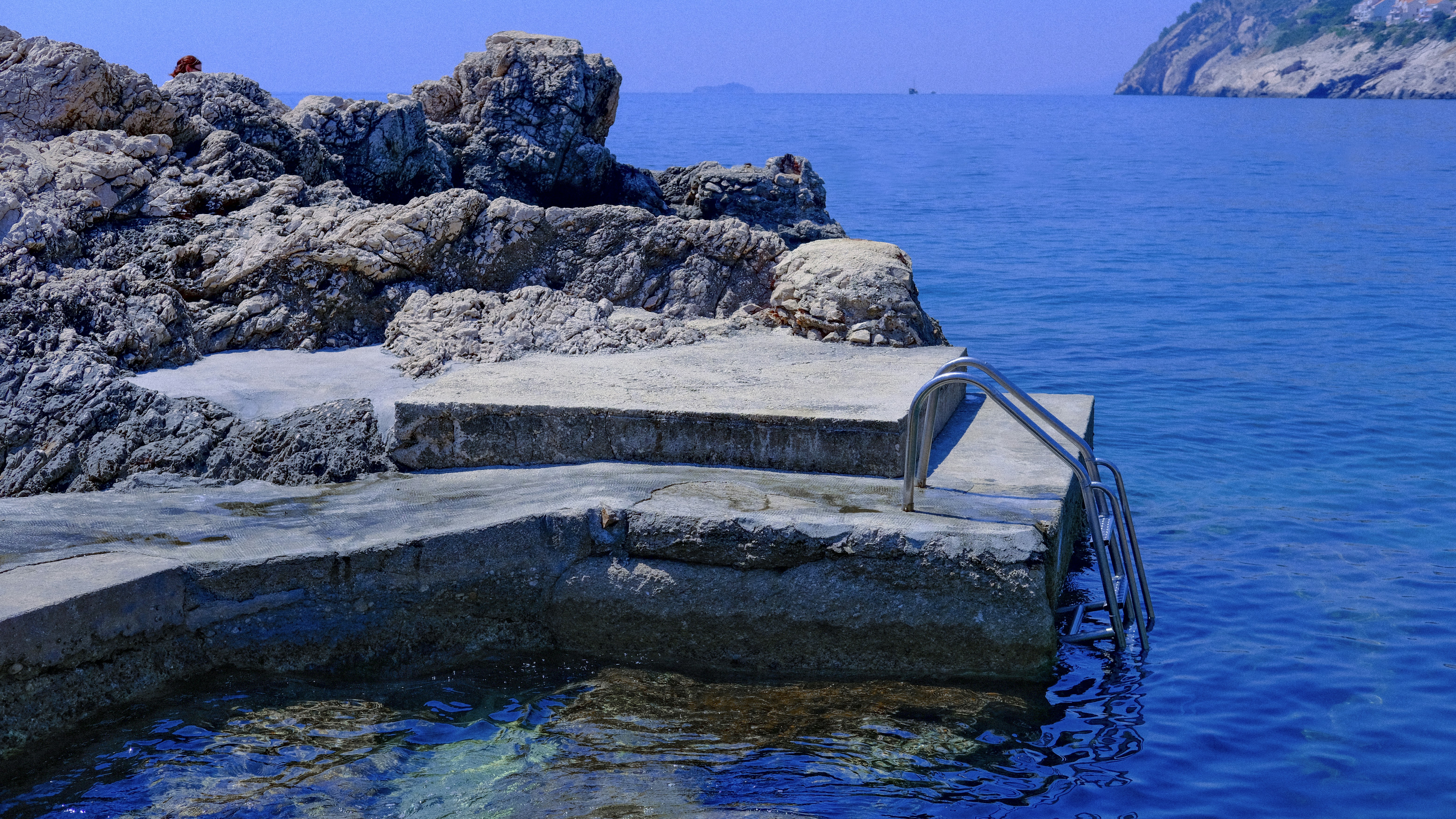 Rocky coastline with a concrete platform leading into calm blue waters, inviting a tranquil escape. A gentle slope hints at the ocean's depths below.